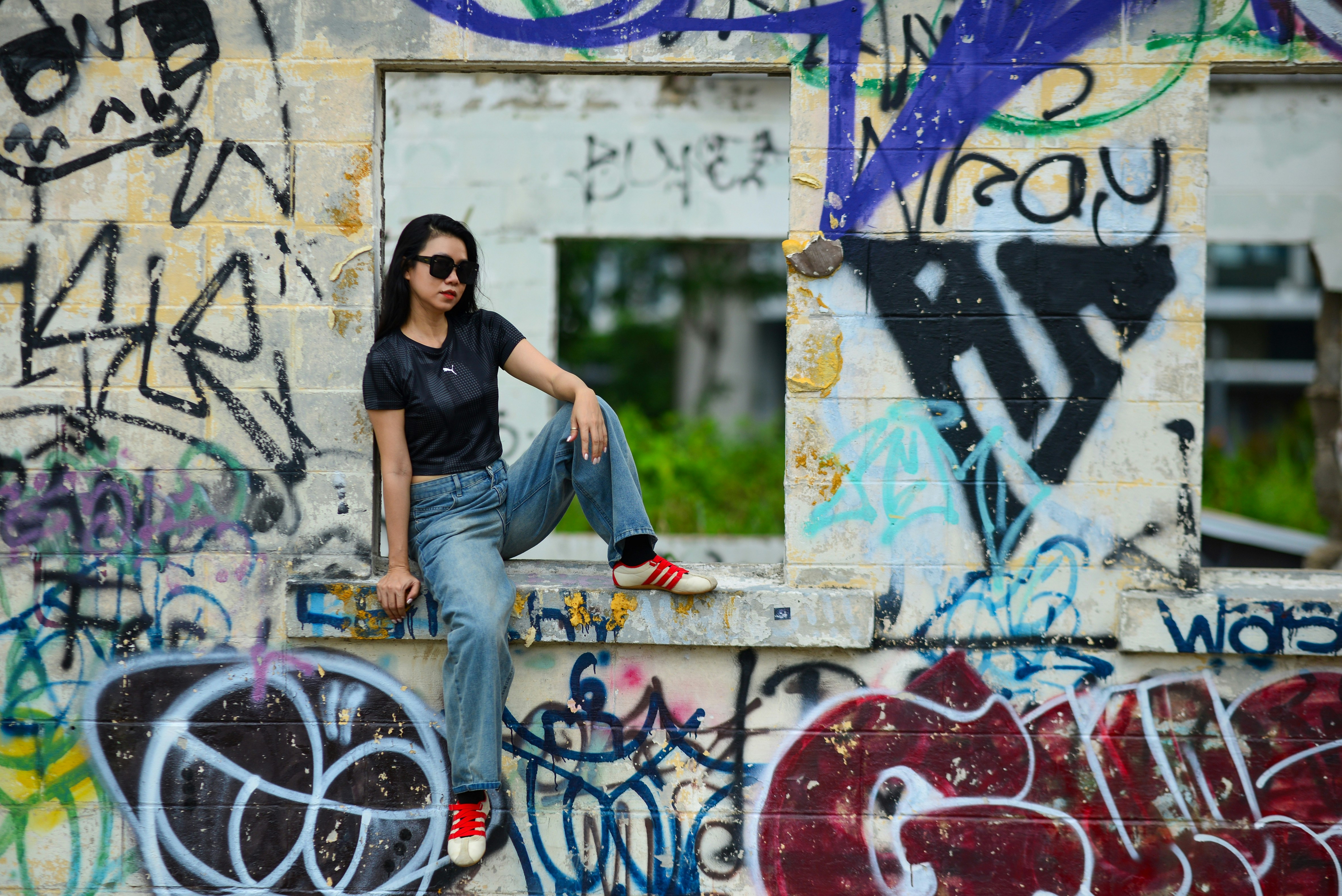 Stylish young Asian woman wearing sunglasses, a black t-shirt, wide leg denim jeans and red and white sneakers poses seated on the ledge of an abandoned graffiti covered concrete building. Urban street fashion mood with vivid spray paint art background, ideal for youth culture, fashion and lifestyle themes.