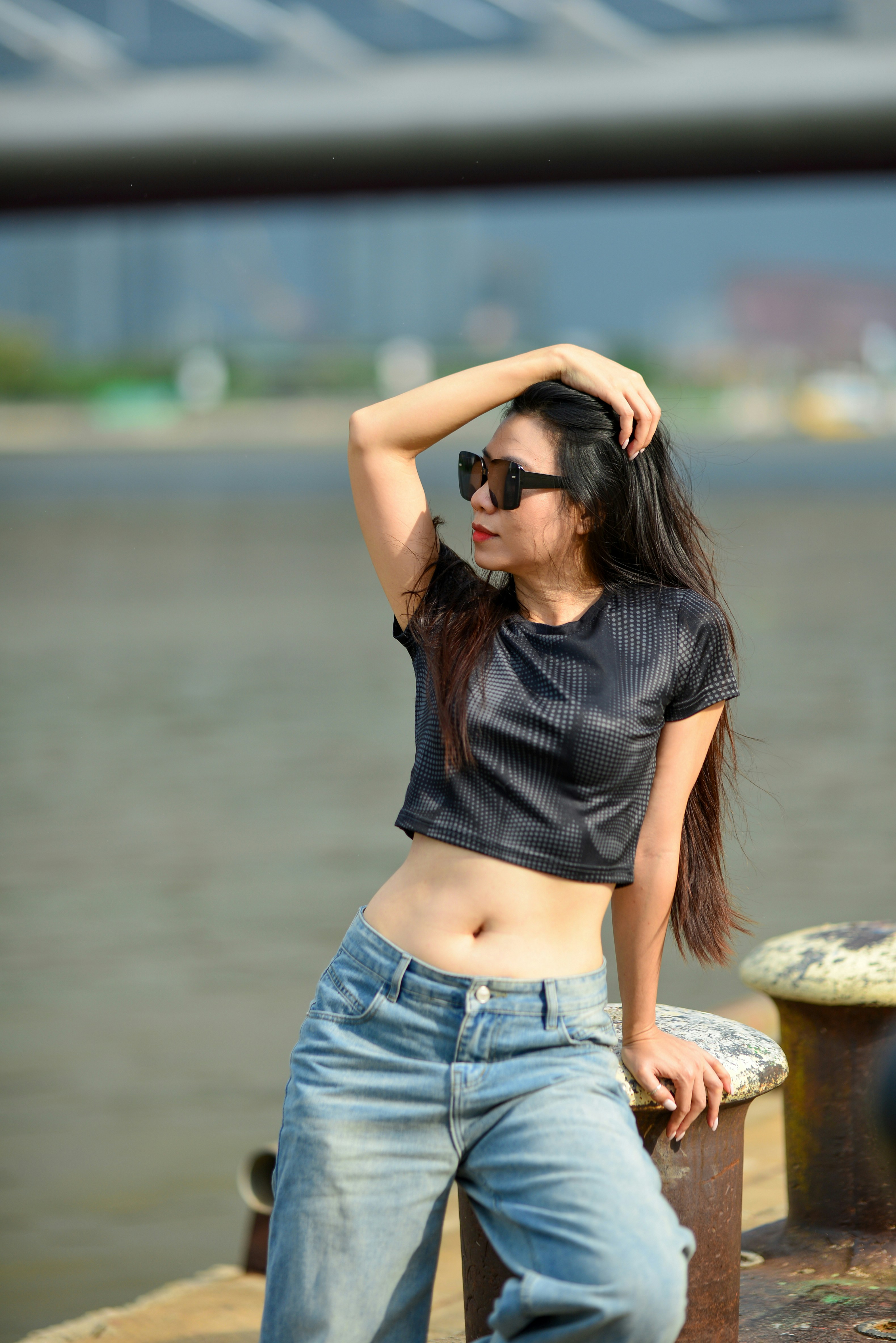 Confident young Asian woman with long dark hair wearing sunglasses, a black crop top and wide-leg denim jeans, leaning against a rusty mooring bollard on a river pier. Soft blurred bridge and city background. Ideal for fashion, urban lifestyle, summer style and streetwear promotion.