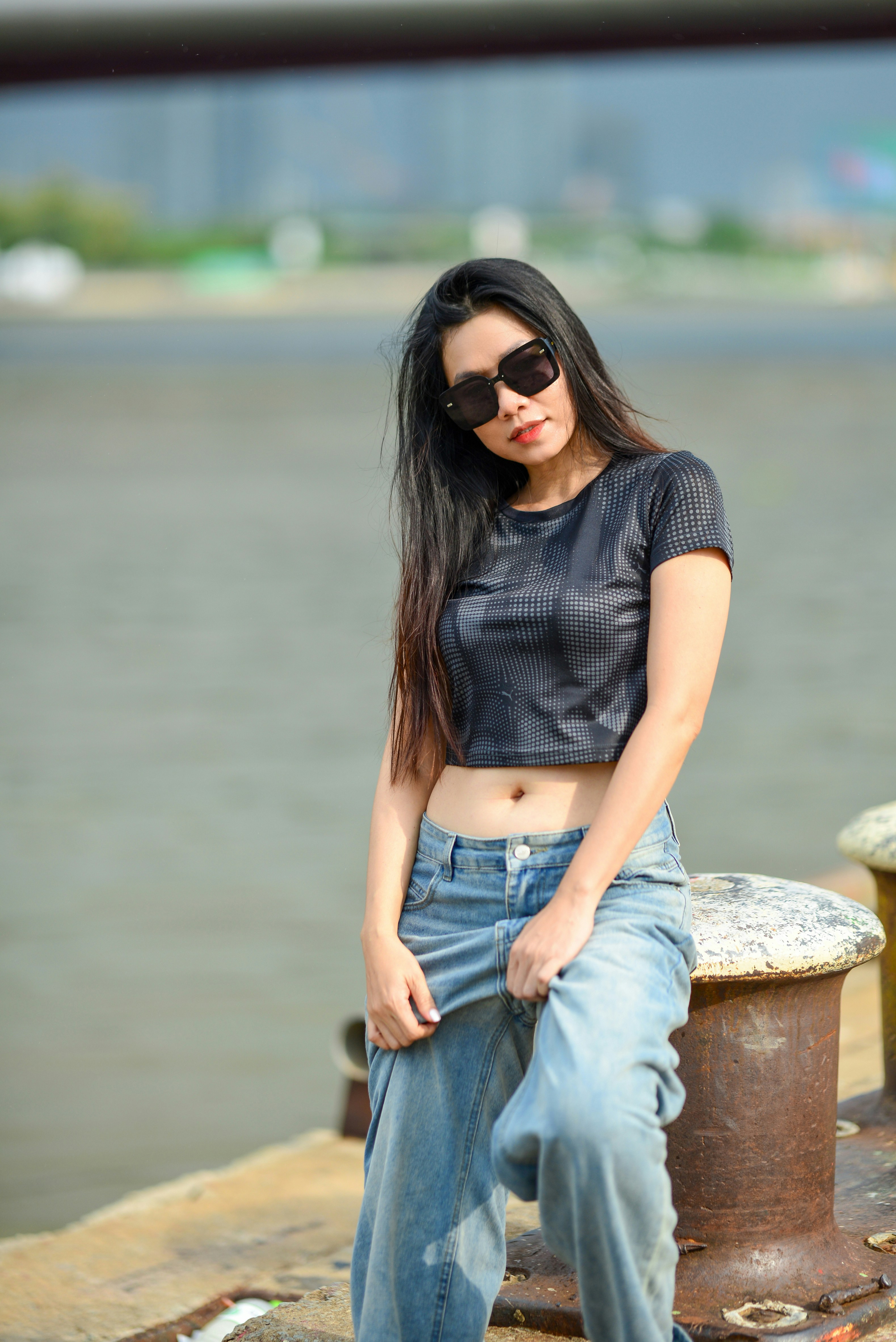 Confident young Asian woman with long dark hair wears oversized sunglasses, a fitted black crop top and loose light blue jeans while leaning against a weathered mooring bollard on a riverside pier. Soft blurred water and bridge in the background. Ideal for urban fashion, streetwear, lifestyle and summer style concepts.