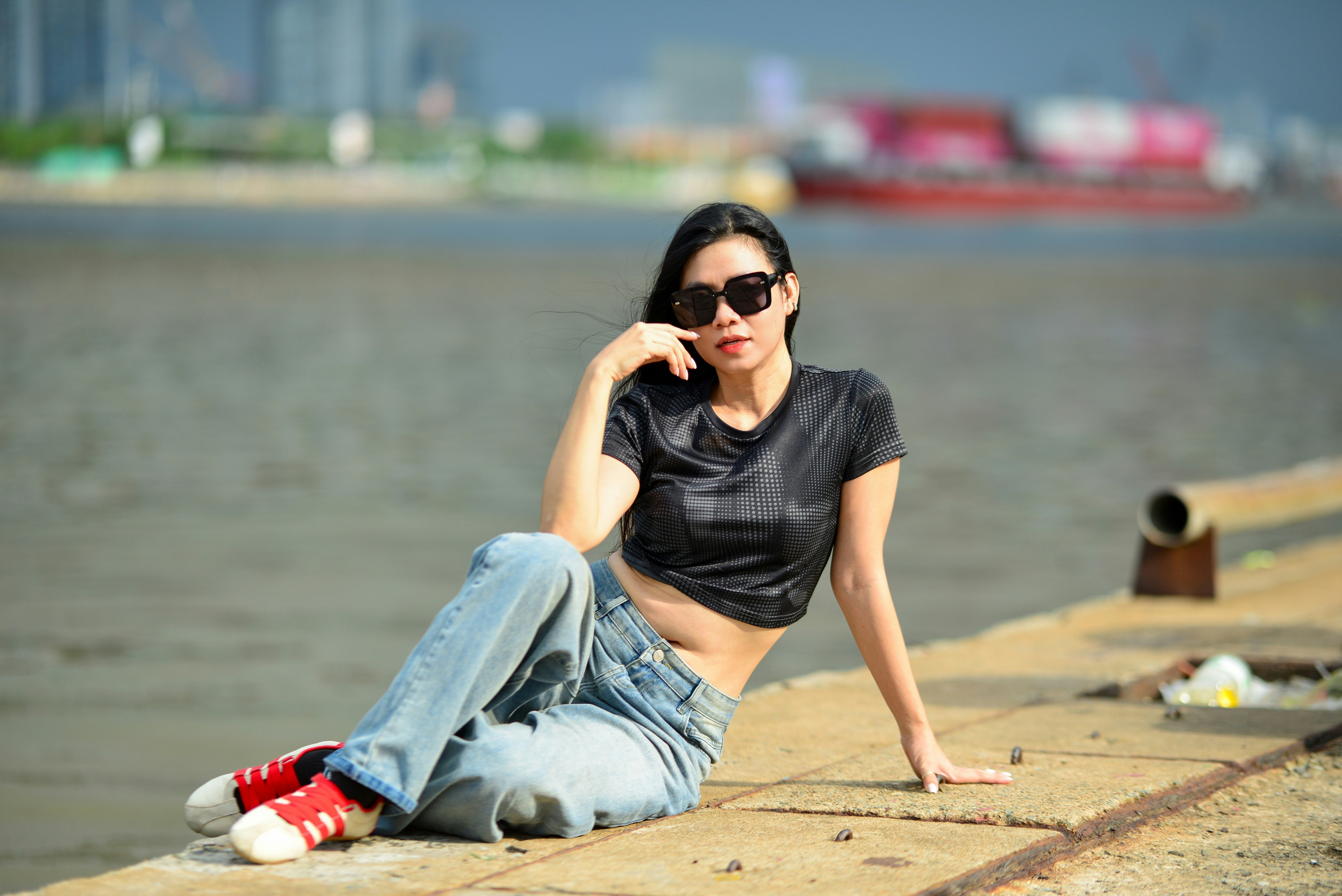 Confident young Asian woman wearing black crop top, wide leg light blue jeans and red sneakers sits on a concrete riverside pier with a cargo barge and city skyline blurred behind her. Urban fashion portrait with strong summer light, useful for lifestyle, streetwear, travel and youth culture concepts.