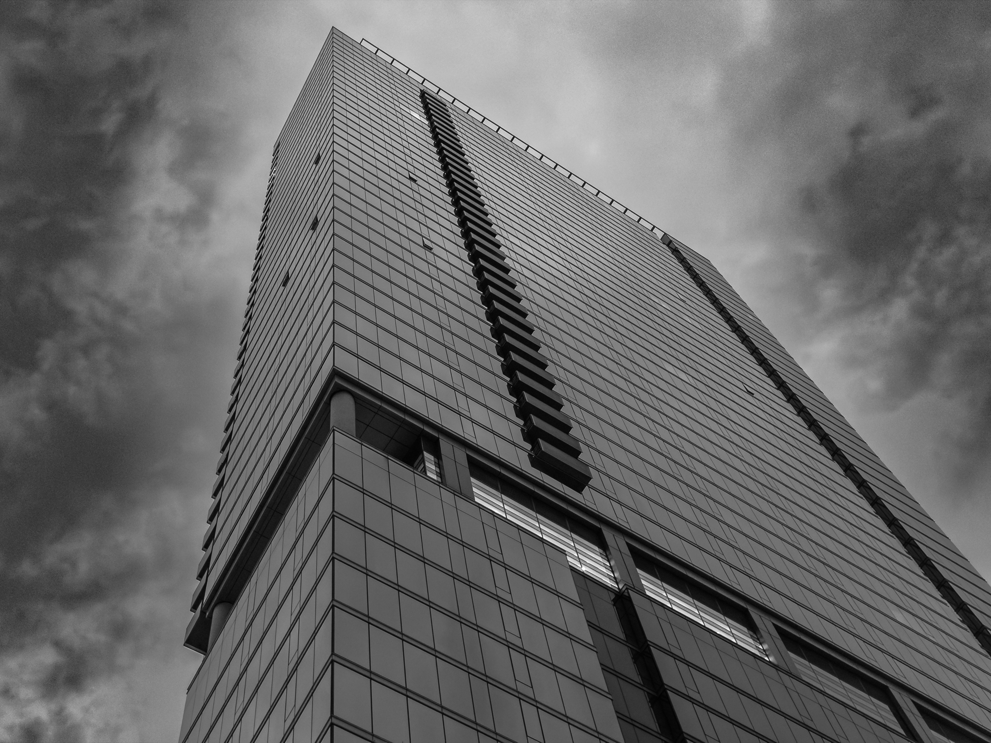 Tall modern skyscraper rising against a cloudy sky, captured from a low angle with bold geometric lines and a monochrome palette.