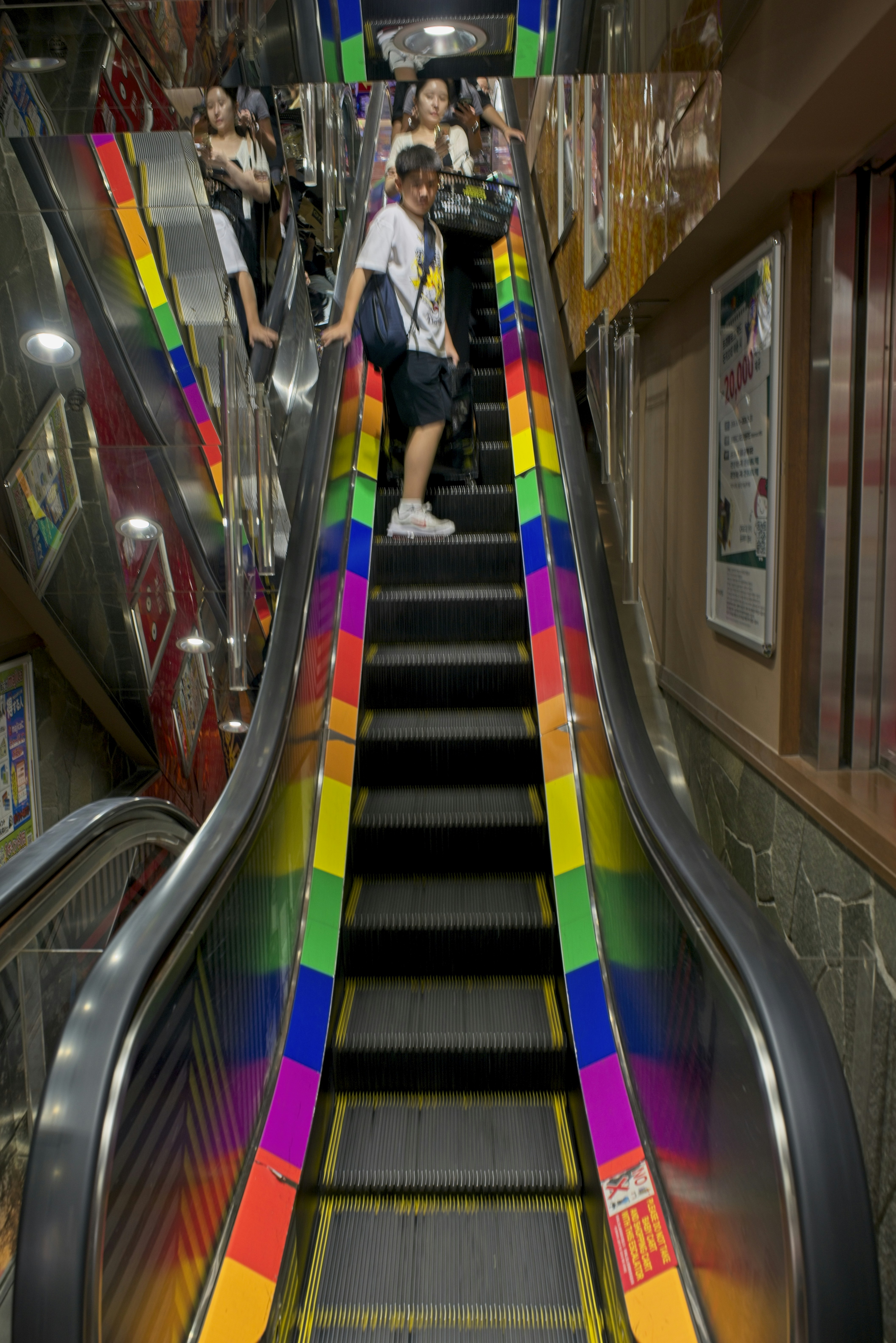 Colorful escalator in a busy indoor space with commuters ascending; glossy rainbow rails mirror the surroundings.