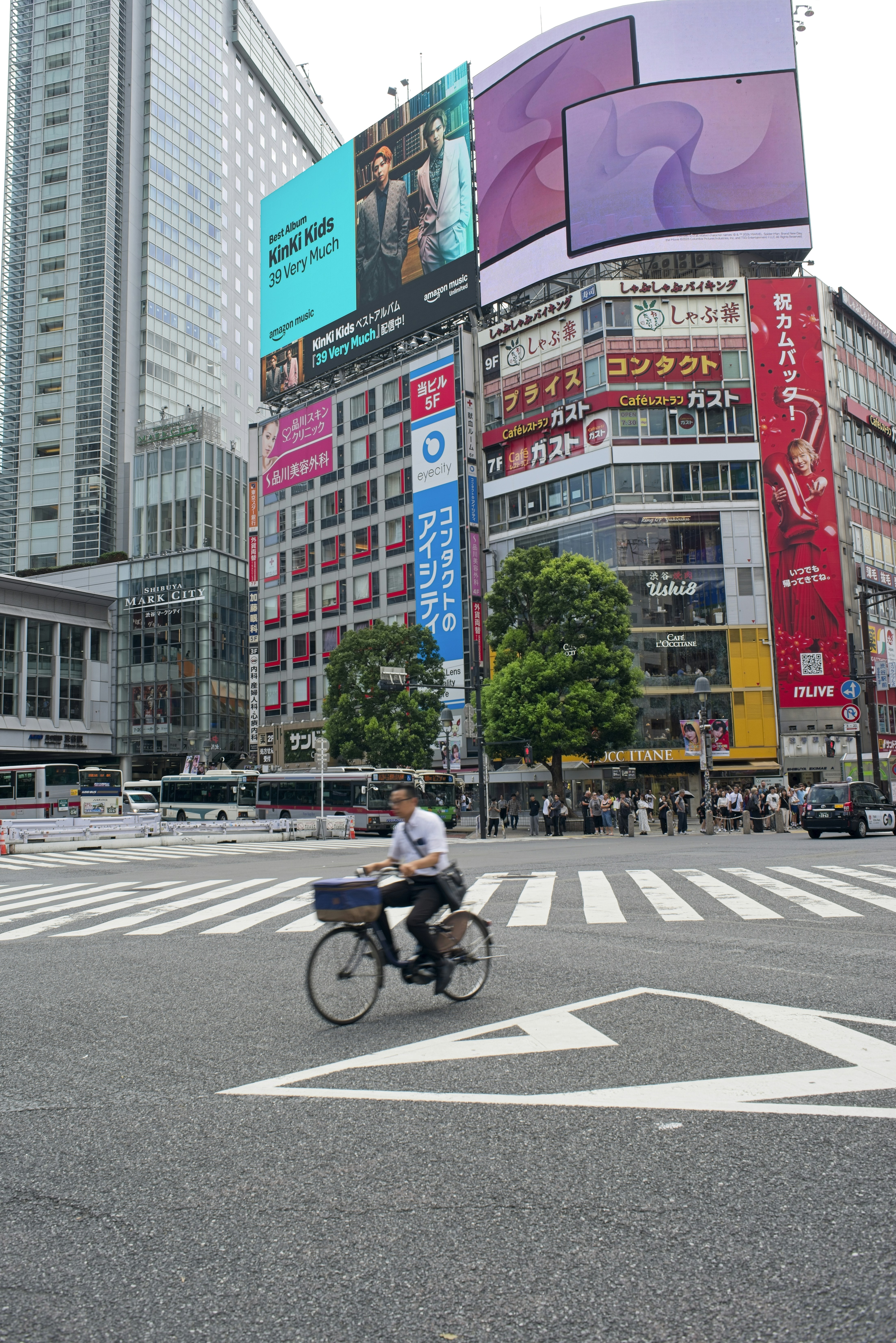 A cyclist crosses the street near Shibuya's iconic intersection, framed by towering buildings covered in vibrant advertisements and digital billboards. Colorful signage promotes restaurants, cosmetics, and pop culture, including a large "SHINE" idol group ad and glowing screens displaying abstract visuals and cityscapes. Cars pause at the crossing while pedestrians gather in the background near storefronts like L'Occitane and Ushi8. Under a hazy grey July sky, the scene captures Shibuya's signature blend of commerce, motion, and sensory overload in one bustling street-level snapshot.
