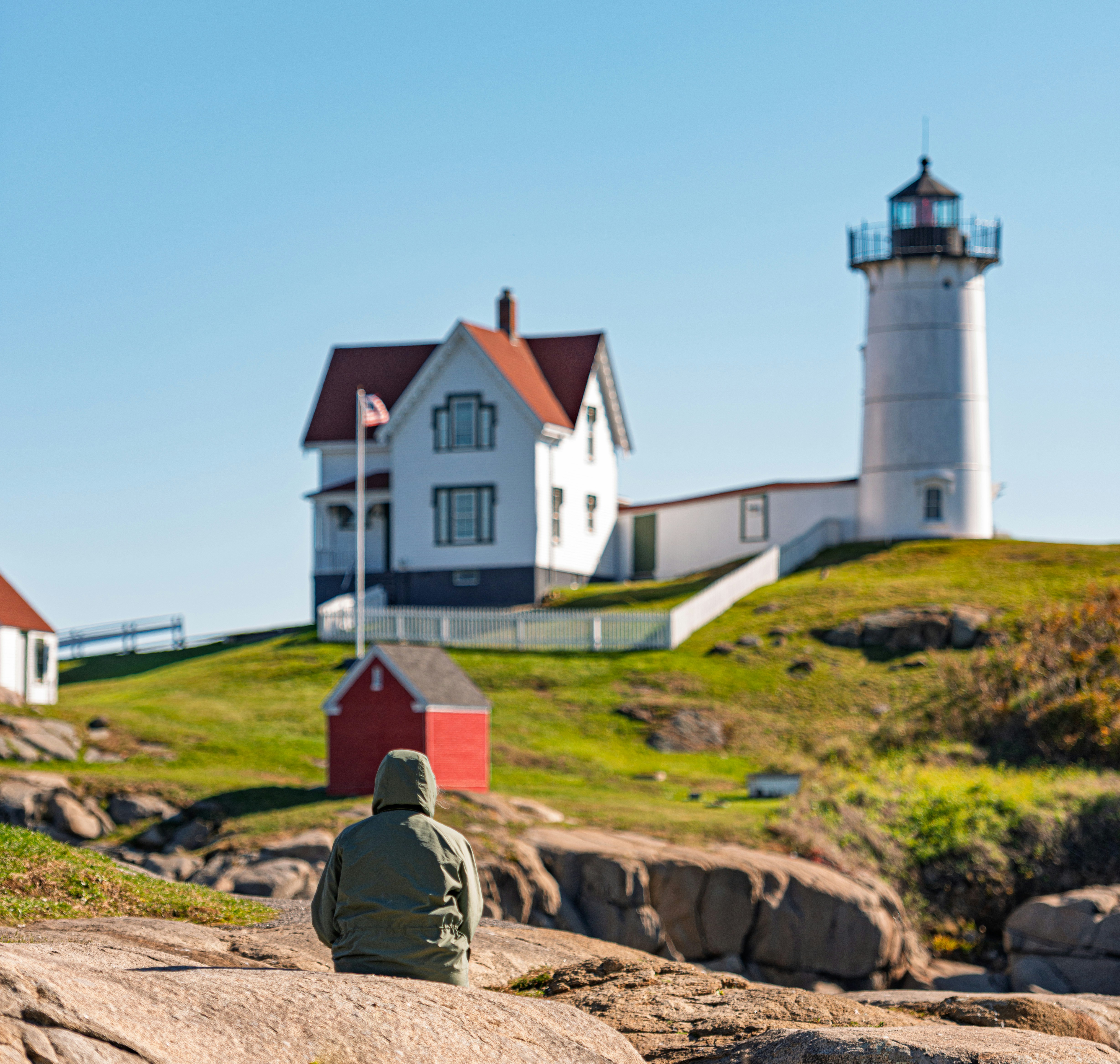 A solitary figure sits on rocky shore facing a white lighthouse and coastal home under a clear blue sky.