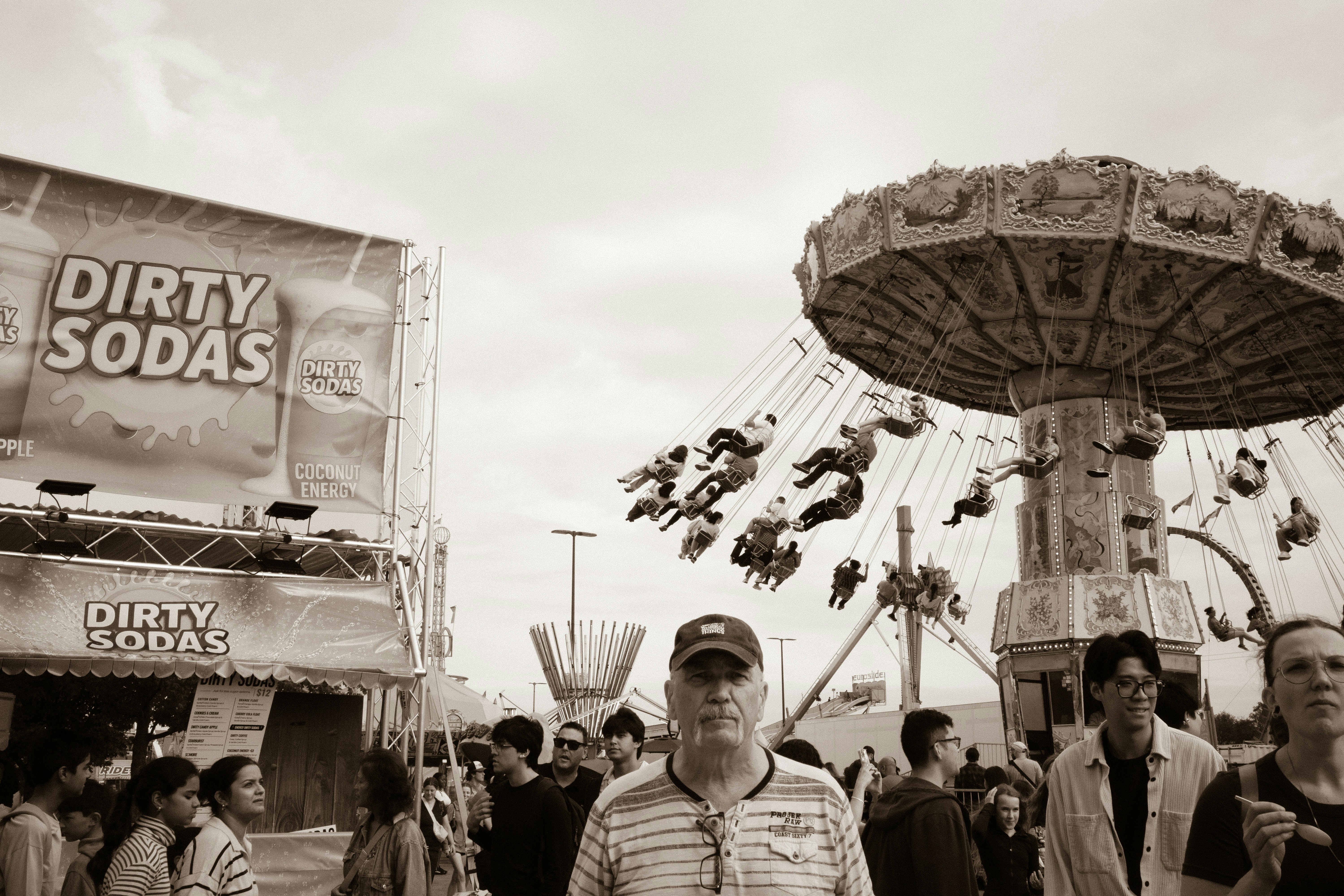 Crowd gathers at a vintage fair with a spinning carousel to the right and a striped vendor stall to the left, creating a nostalgic street-scene composition.