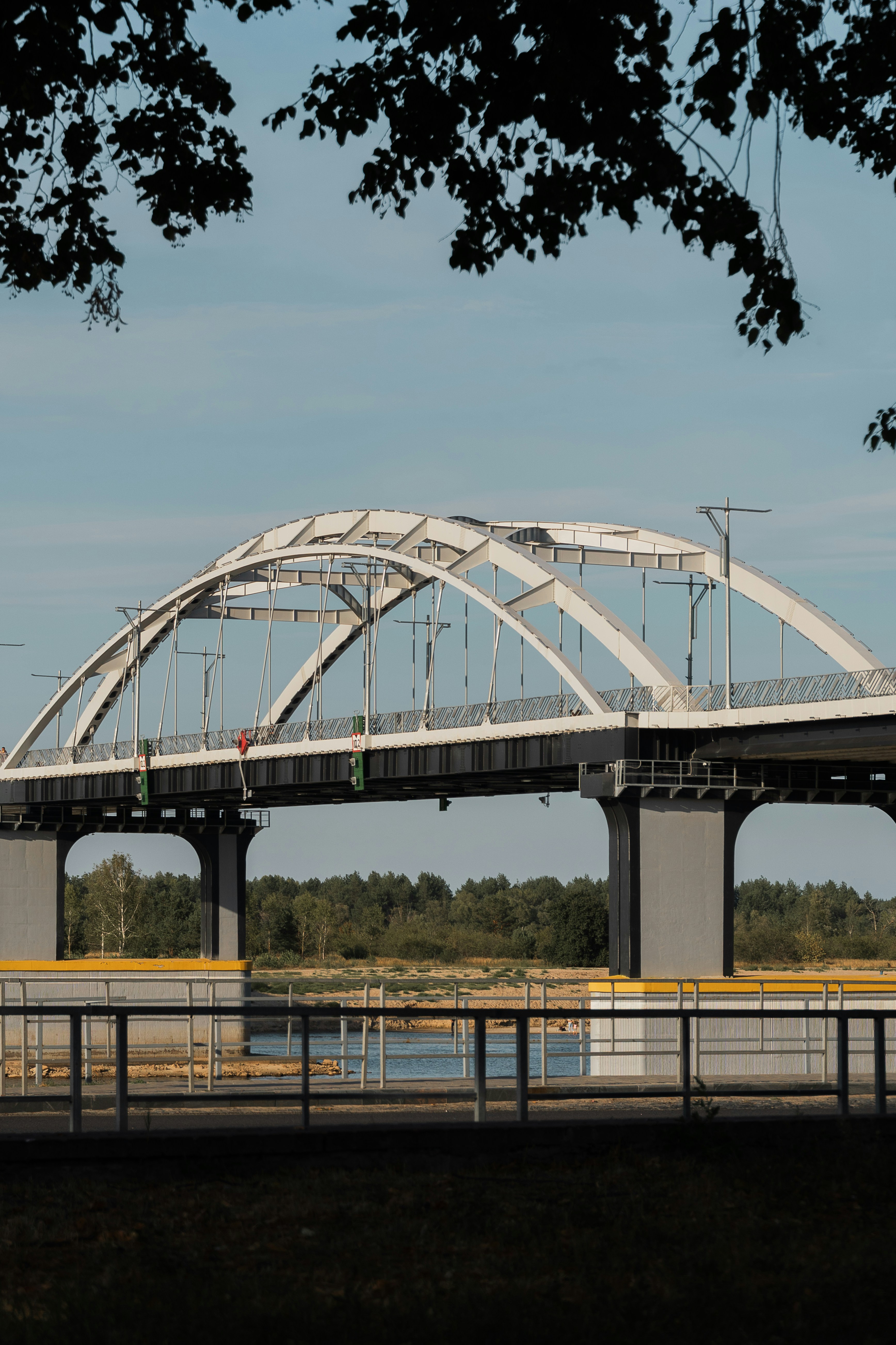 Bridge with white arches spans a calm river under a blue sky; foreground features railing and water reflections, with distant tree line. Aerial perspective emphasizes structure and open space. This is a photography capture of a bridge, not a logo or illustration.