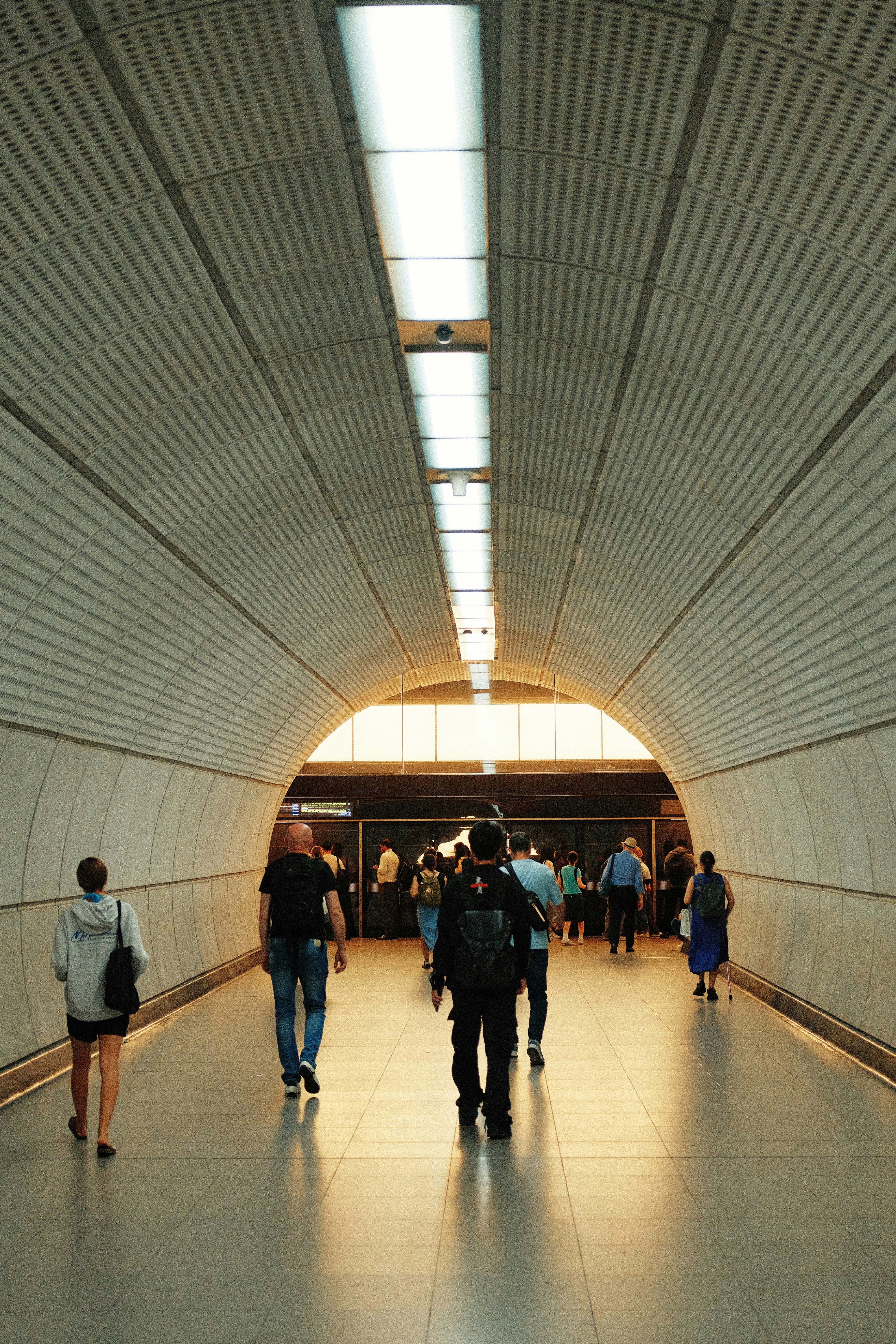 Commuters walk through a wide, arched subway tunnel toward a bright exit. The scene emphasizes symmetry and movement within an industrial, curved environment.