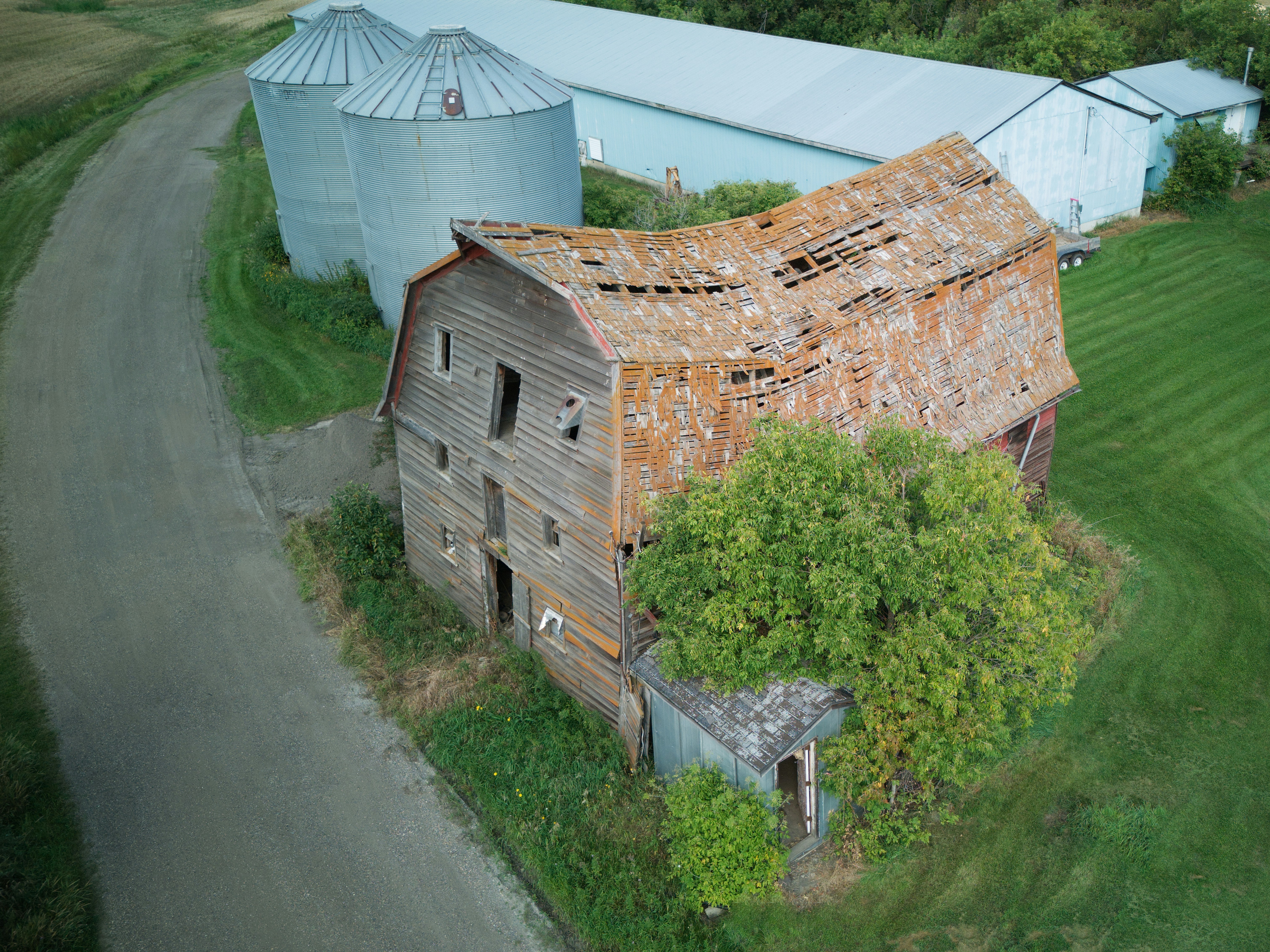 A weathered, multi-story barn with a collapsed roof sits beside silos and a curved driveway, framed by trees and a tidy field.