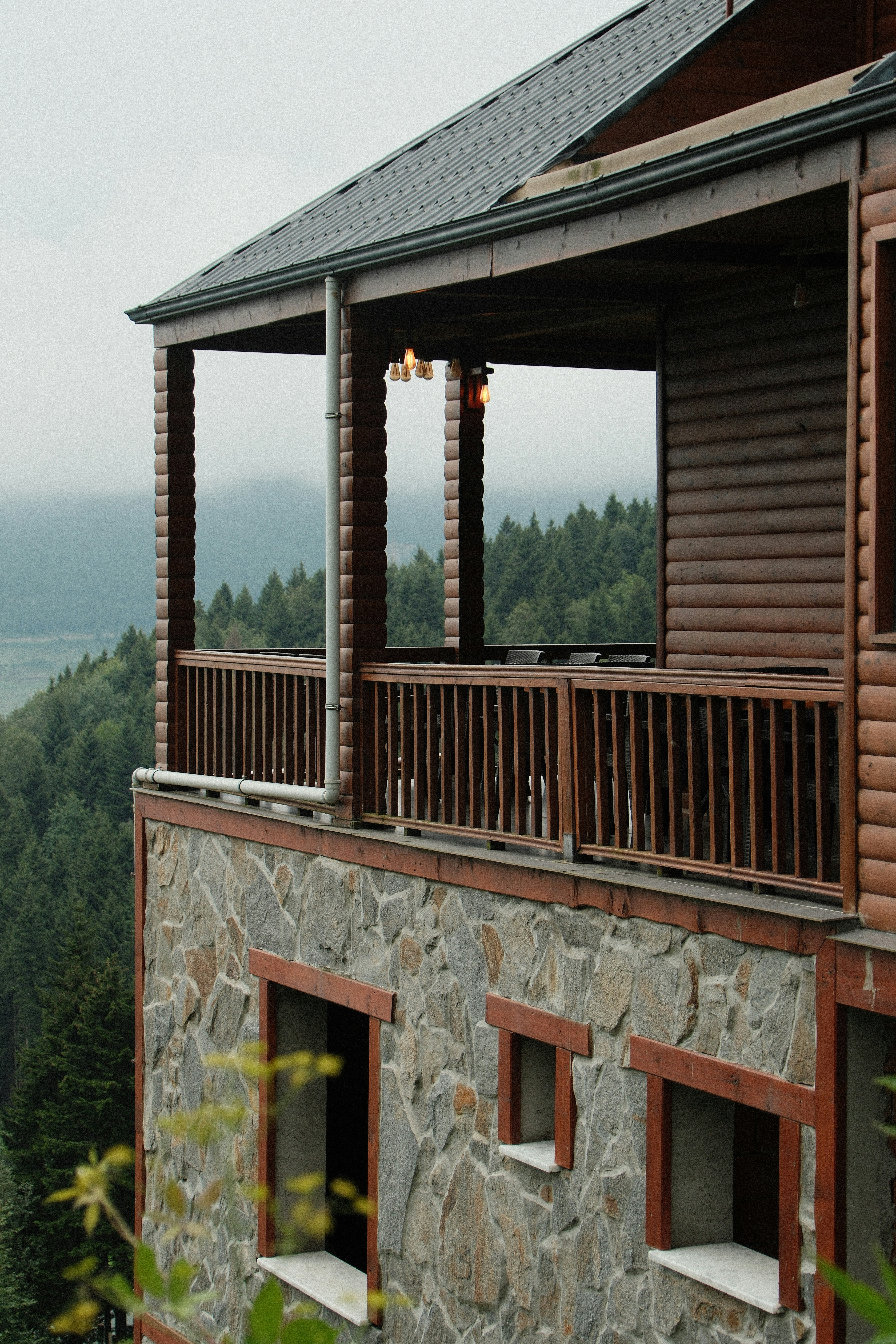 A stone-and-wood cabin with a wraparound balcony sits along a forested hillside, framed by long vertical posts and a railing, overlooking distant treetops and misty mountains.