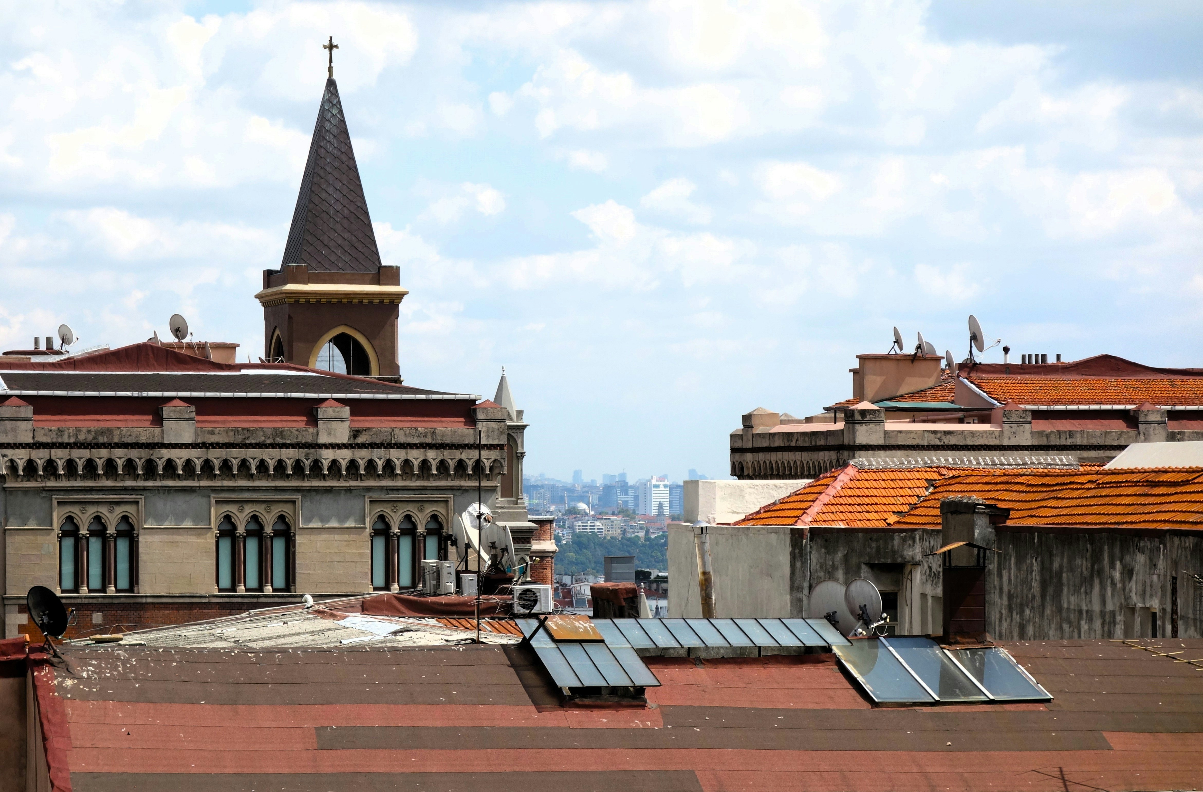 Rooftops with a clock-tower, solar panels and a distant city skyline under a bright, partly cloudy sky.