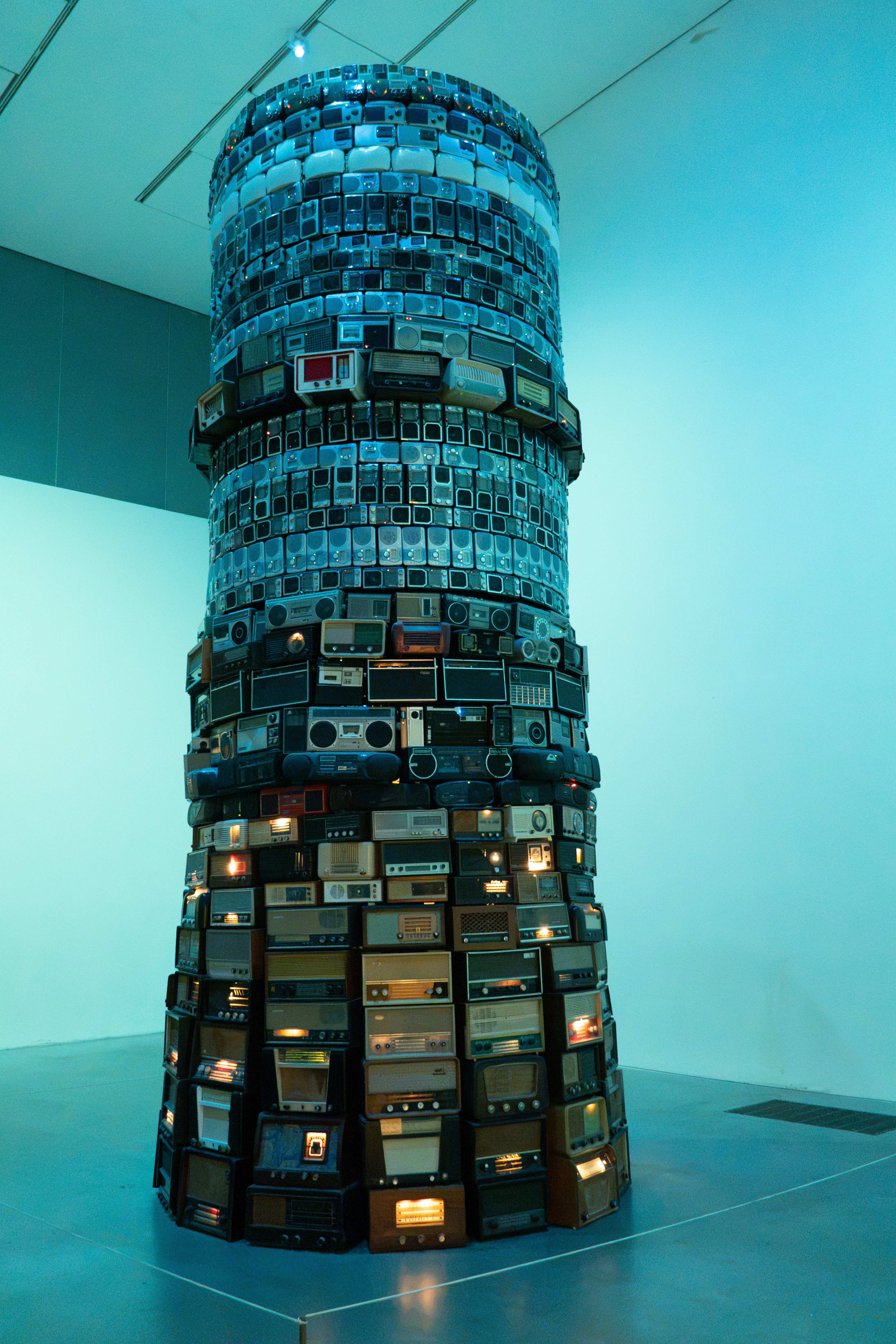 A cylindrical column composed of stacked vintage radios rises from the gallery floor, set against a teal backdrop and softly lit ceiling; the installation doubles as a sculptural archive. This piece is a photographic capture of the exhibit.