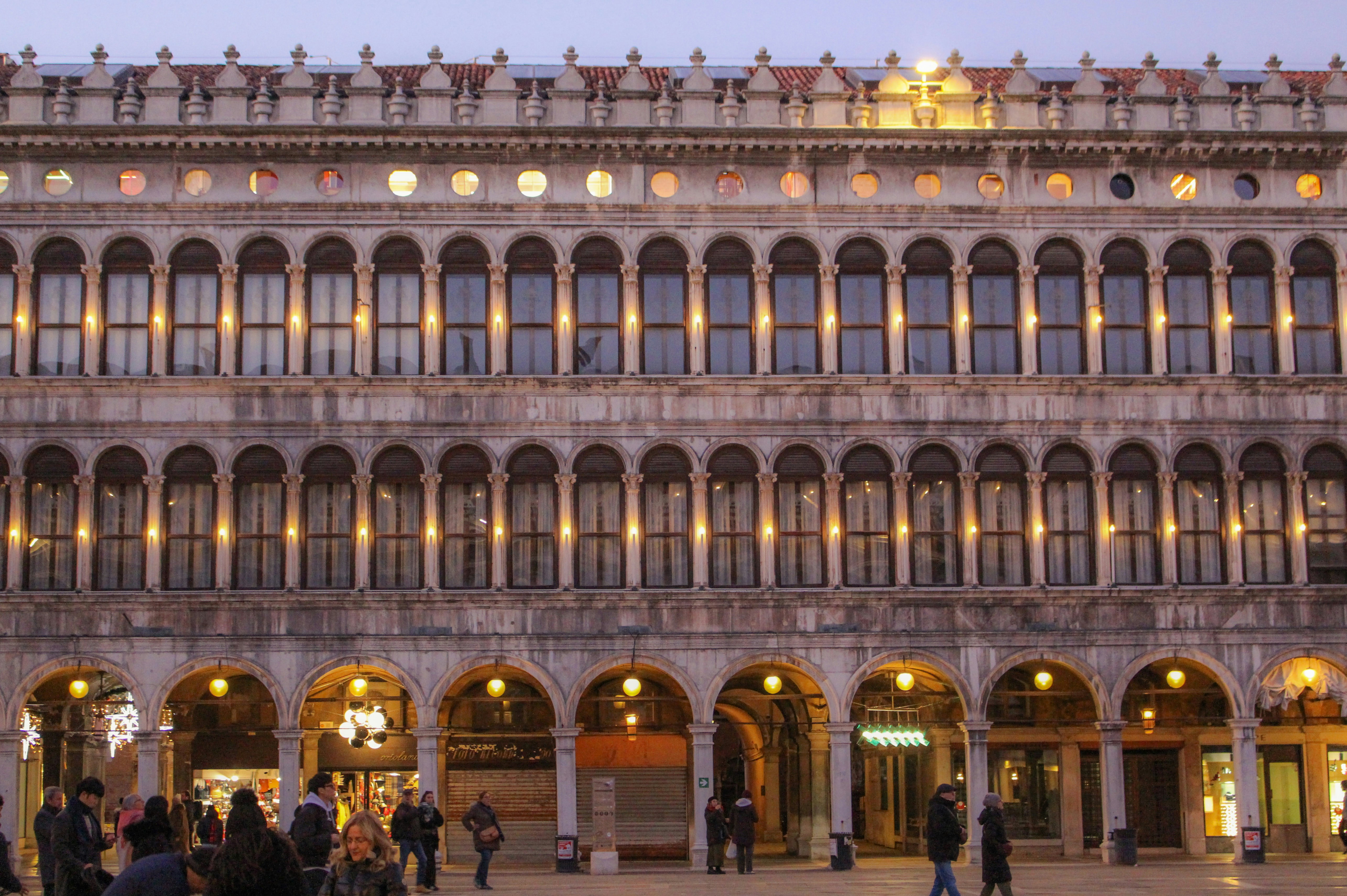 Evening view of a grand arched arcade with illuminated columns and crowds at the ground level, highlighting the repetitive arches and warm glow of streetlights.