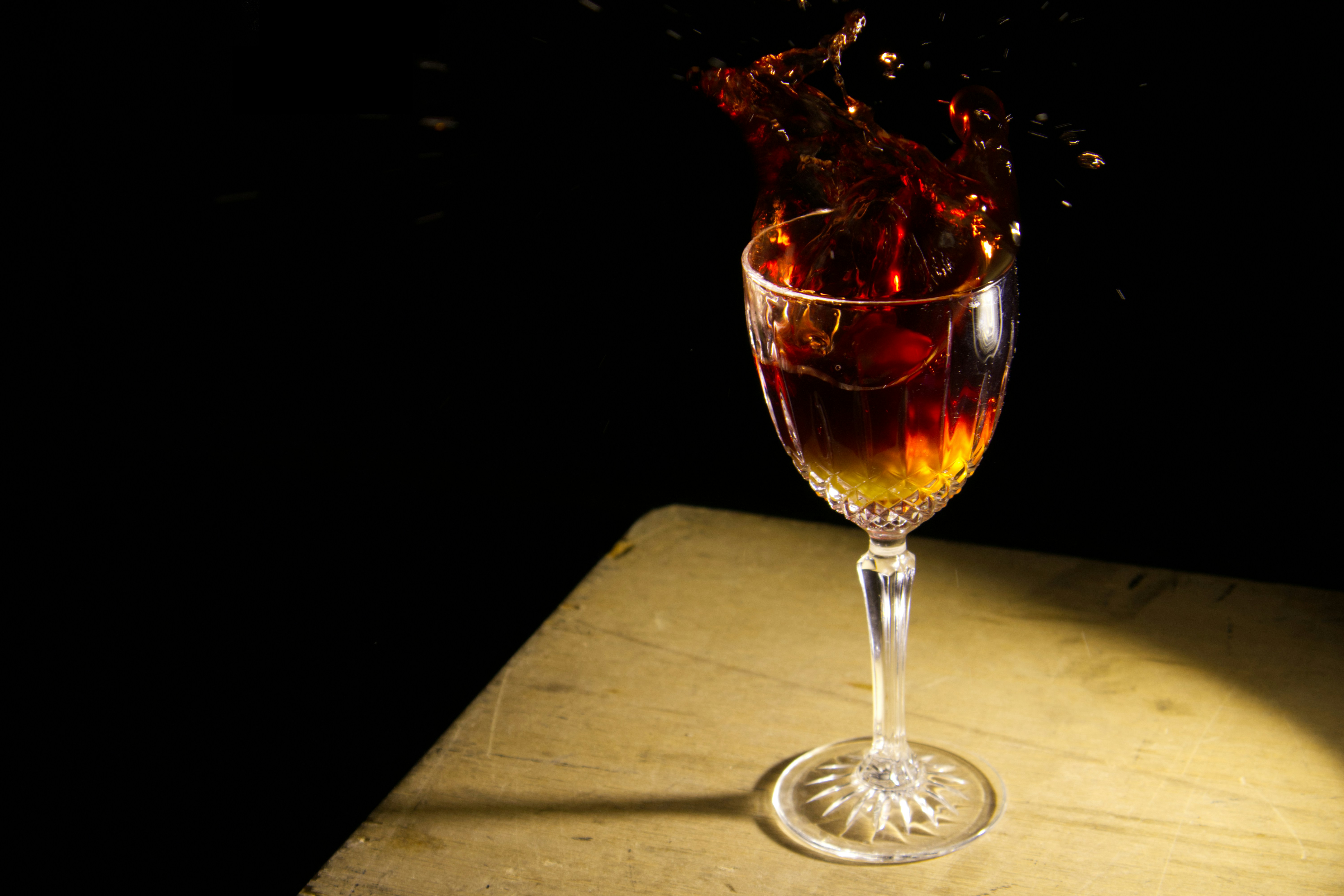 A crystal wine glass sits on a wooden surface as a vivid red liquid splashes into it, captured against a dark background. This is a still-life photograph.