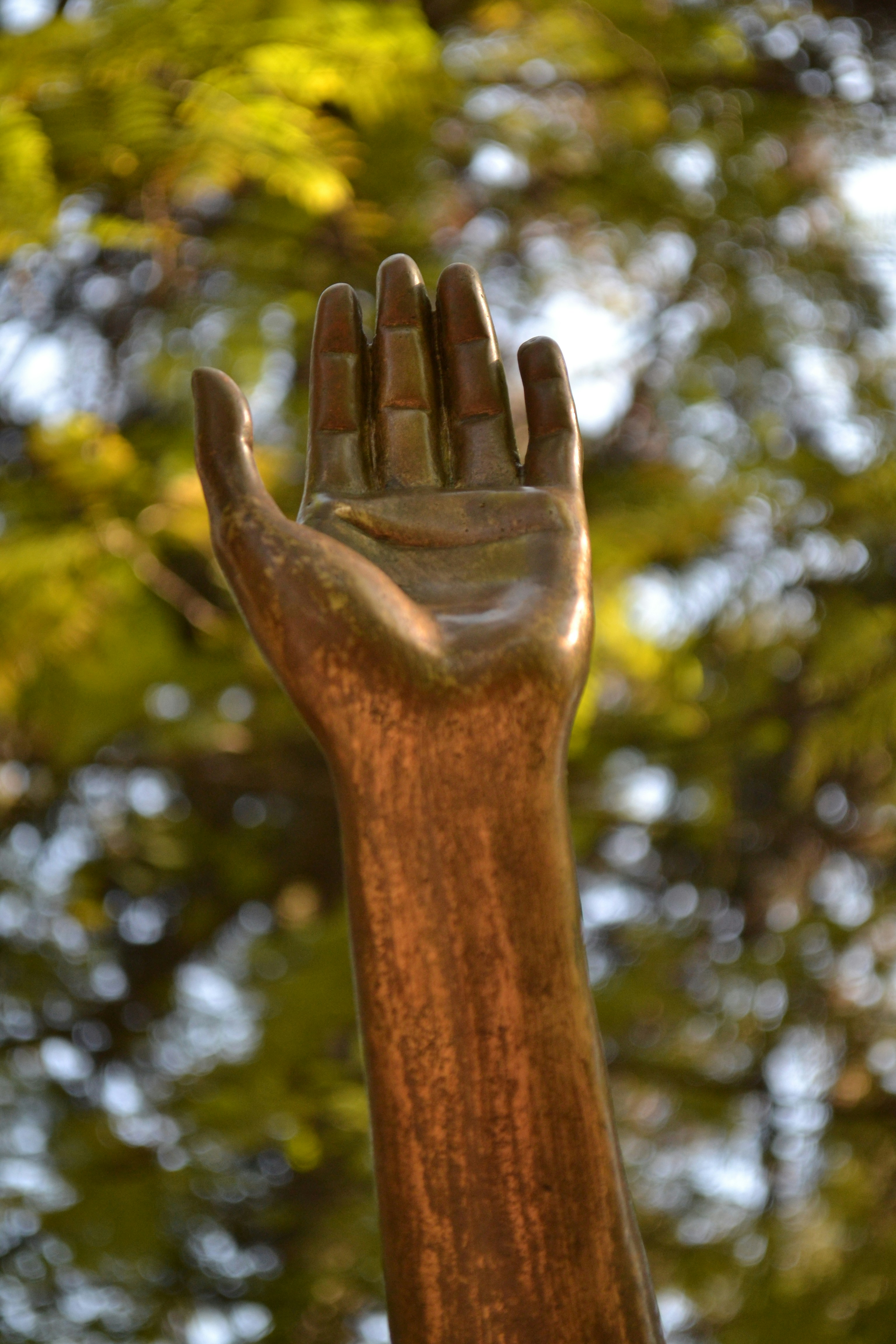 Bronze hand sculpture raised upward against a blurred autumnal background, emphasizing form and patina. The image captures the statue's texture and vertical lines, with the sculpture serving as the focal point.