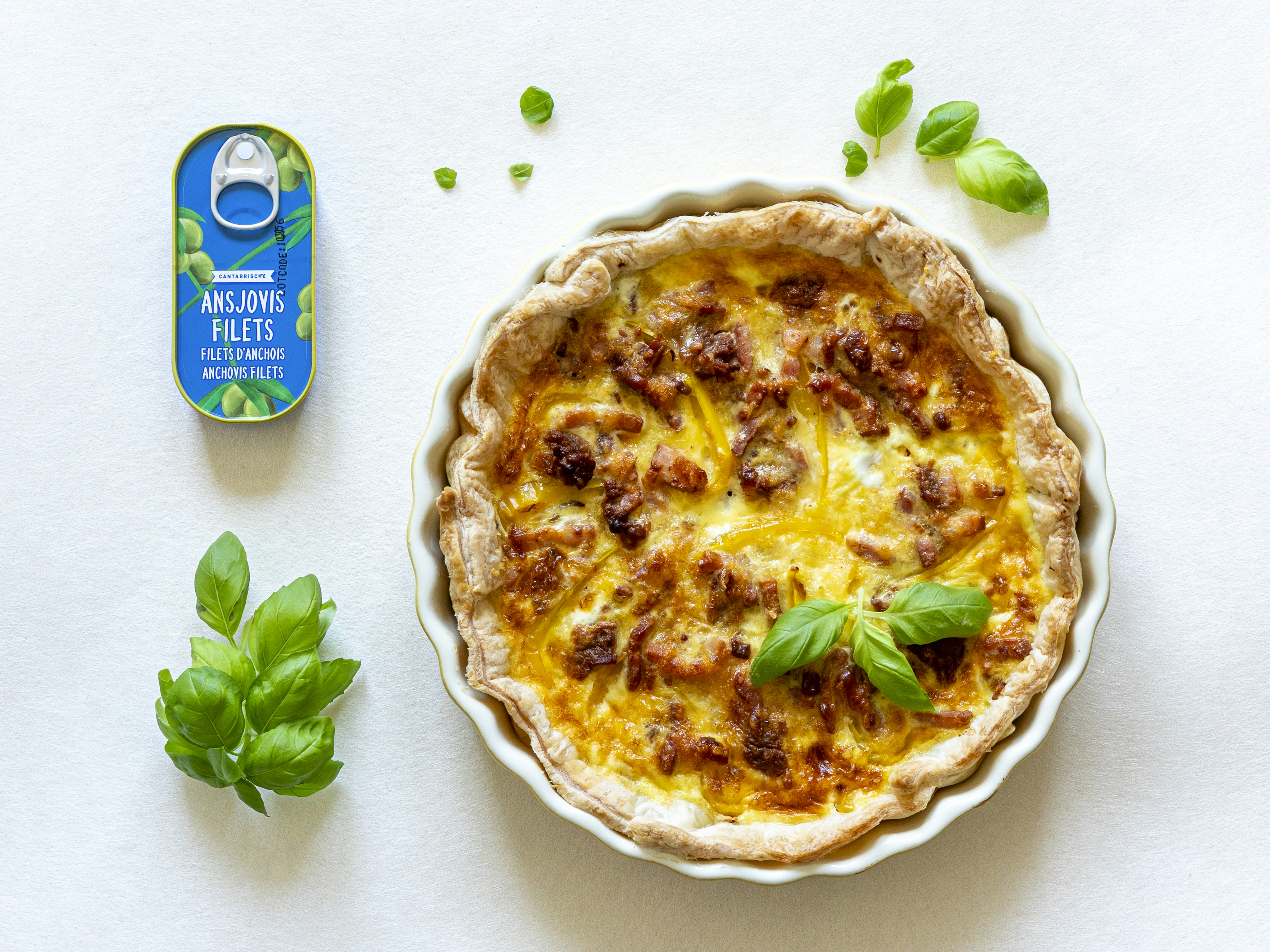 Savory quiche cooling on a pale surface with a small can of tuna-style pâté and fresh basil leaves nearby.
