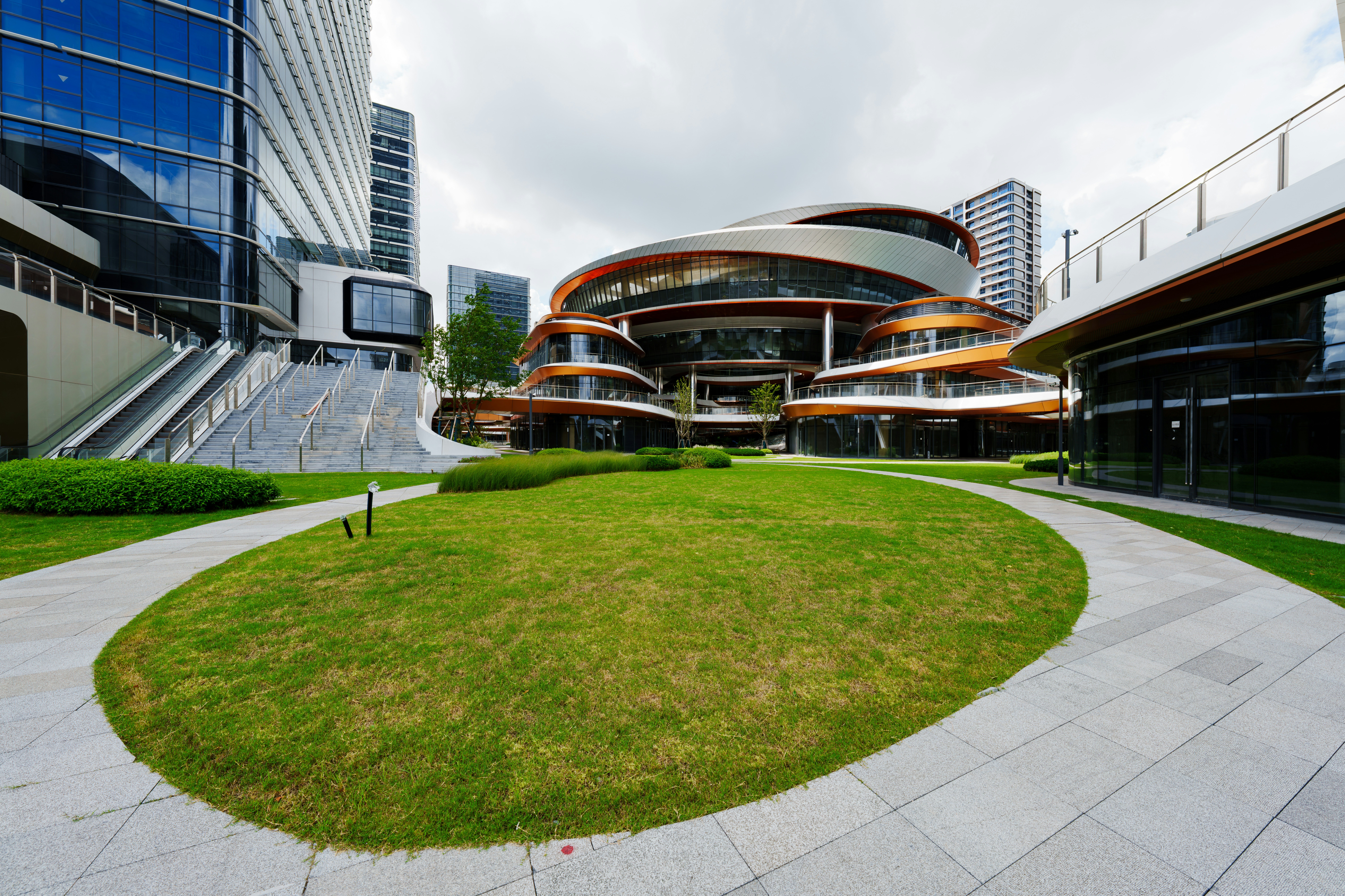 Urban courtyard with a large circular grassy patch surrounded by light stone paths, flanked by futuristic, curved orange-and-wood buildings and glass towers.