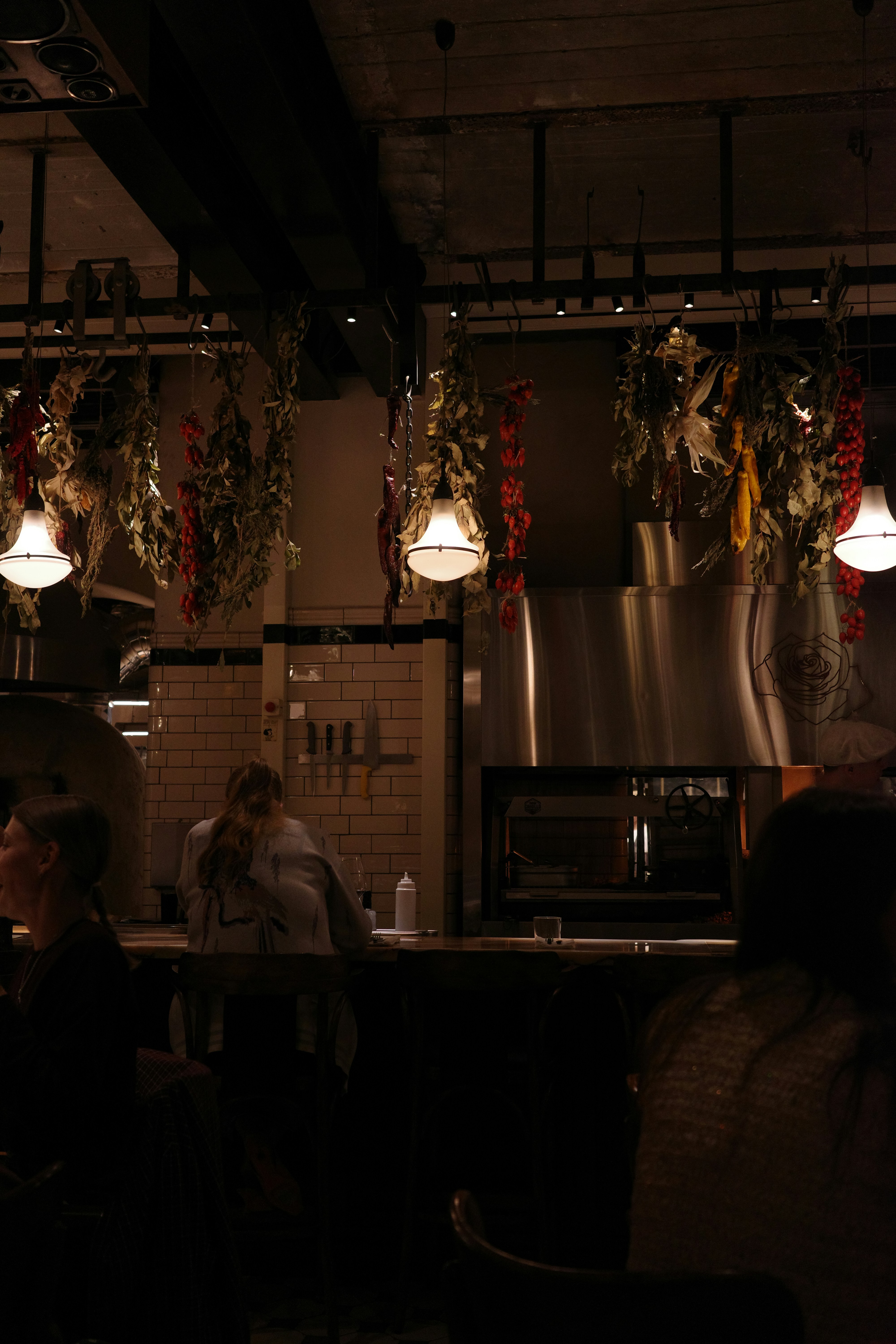Dimly lit restaurant kitchen area with hanging dried herbs and warm pendant lights over a counter; blurred diners in foreground and stainless steel kitchen equipment in back.