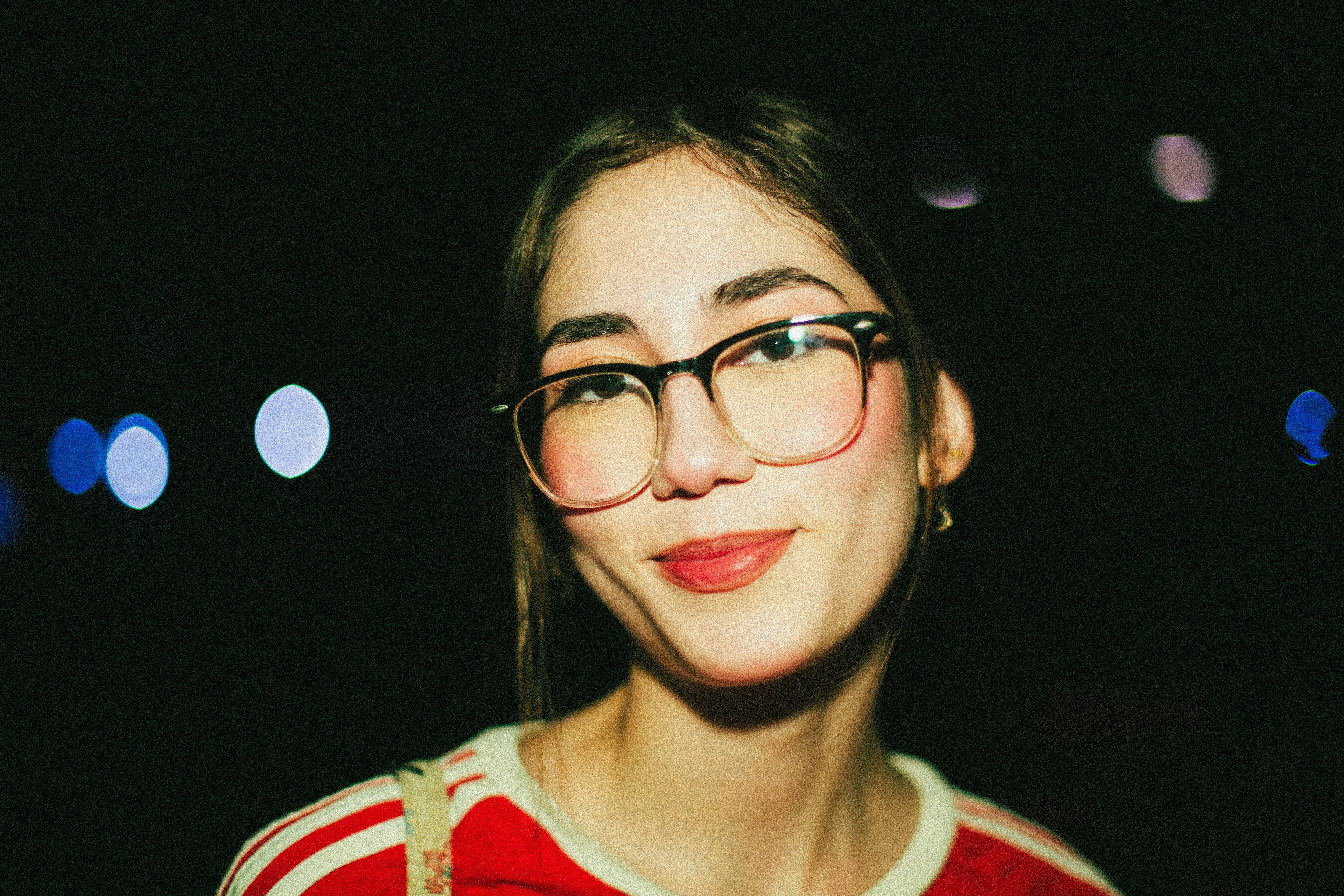 Portrait of a woman wearing glasses at night, soft bokeh lights in the background; a candid, warm-toned head-and-shoulders shot. This is a photographic capture, not a drawing or logo.