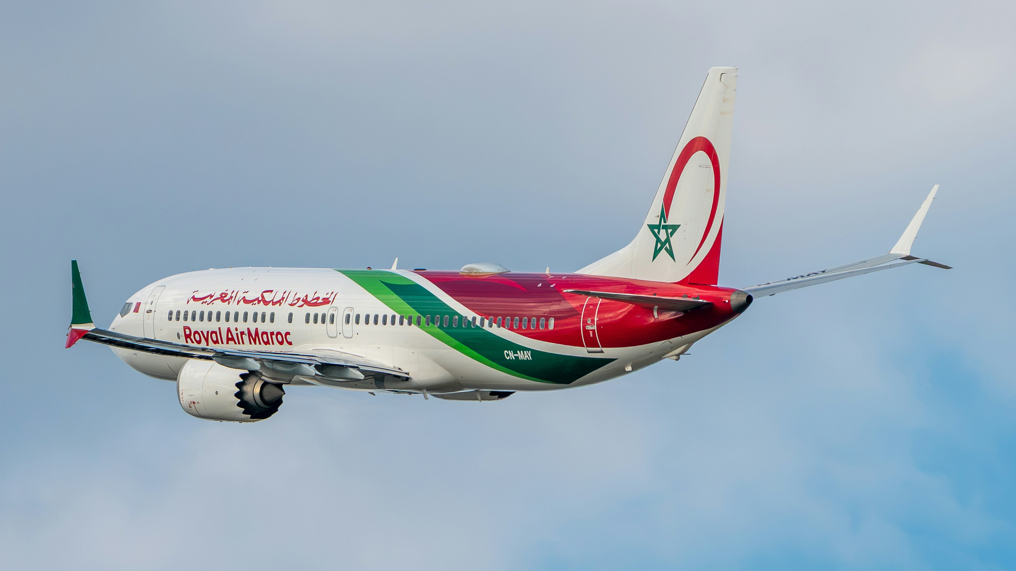 A Royal Air Maroc aircraft in flight with the distinctive green, red, and white livery against a soft blue sky; the image captures the plane mid-ascent with emphasis on branding and silhouette. This is a photograph of a real airline aircraft.