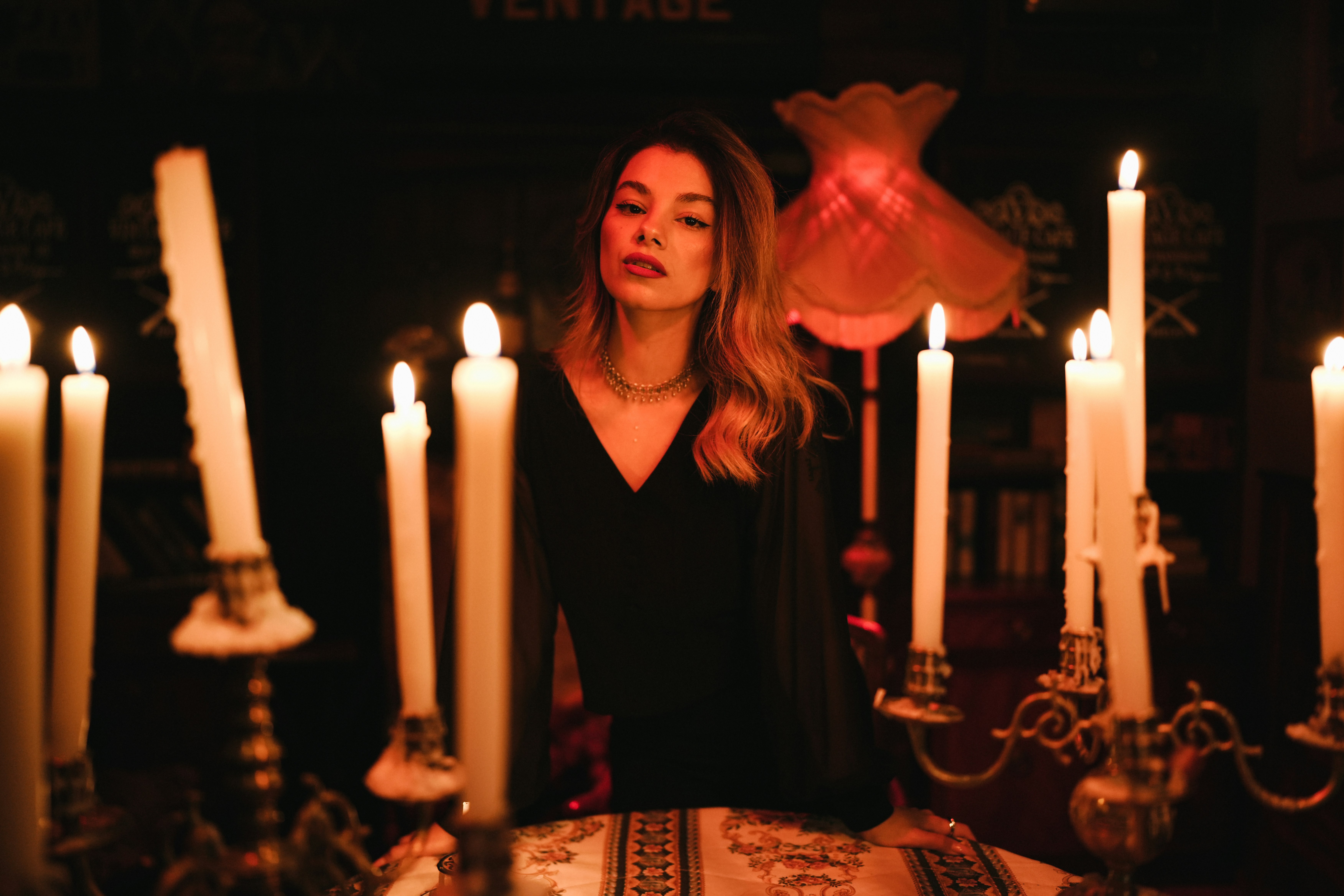 A moody portrait of a woman seated at a candlelit table, surrounded by tall candles and ornate surroundings in a dark restaurant or venue. This is a photographic scene with warm light and atmospheric ambience.