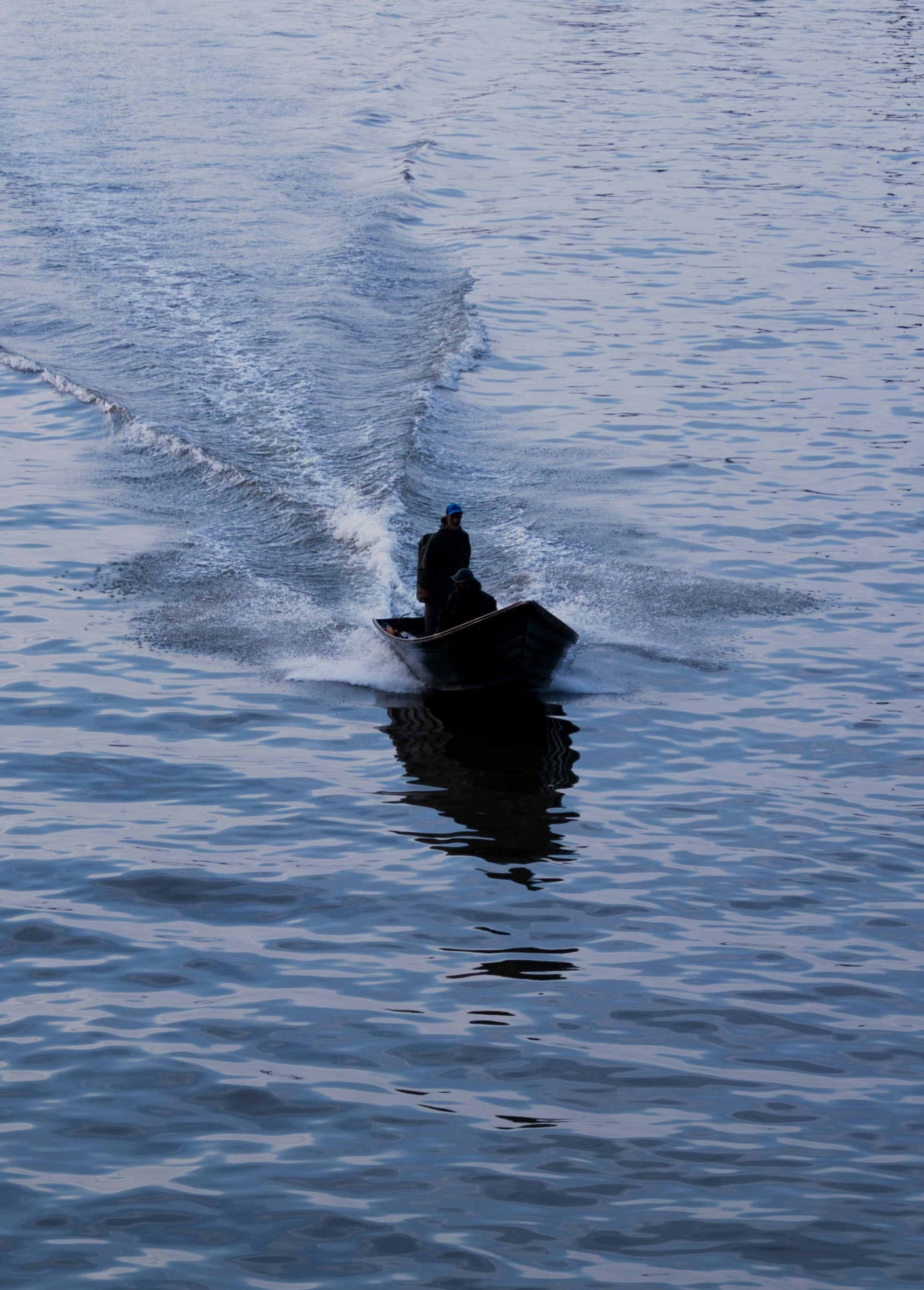 Small motorboat cuts a long wake across calm blue water, two figures visible at the bow with a clear reflection trailing behind. The scene emphasizes motion and contrast between the dark silhouette and tranquil surface.