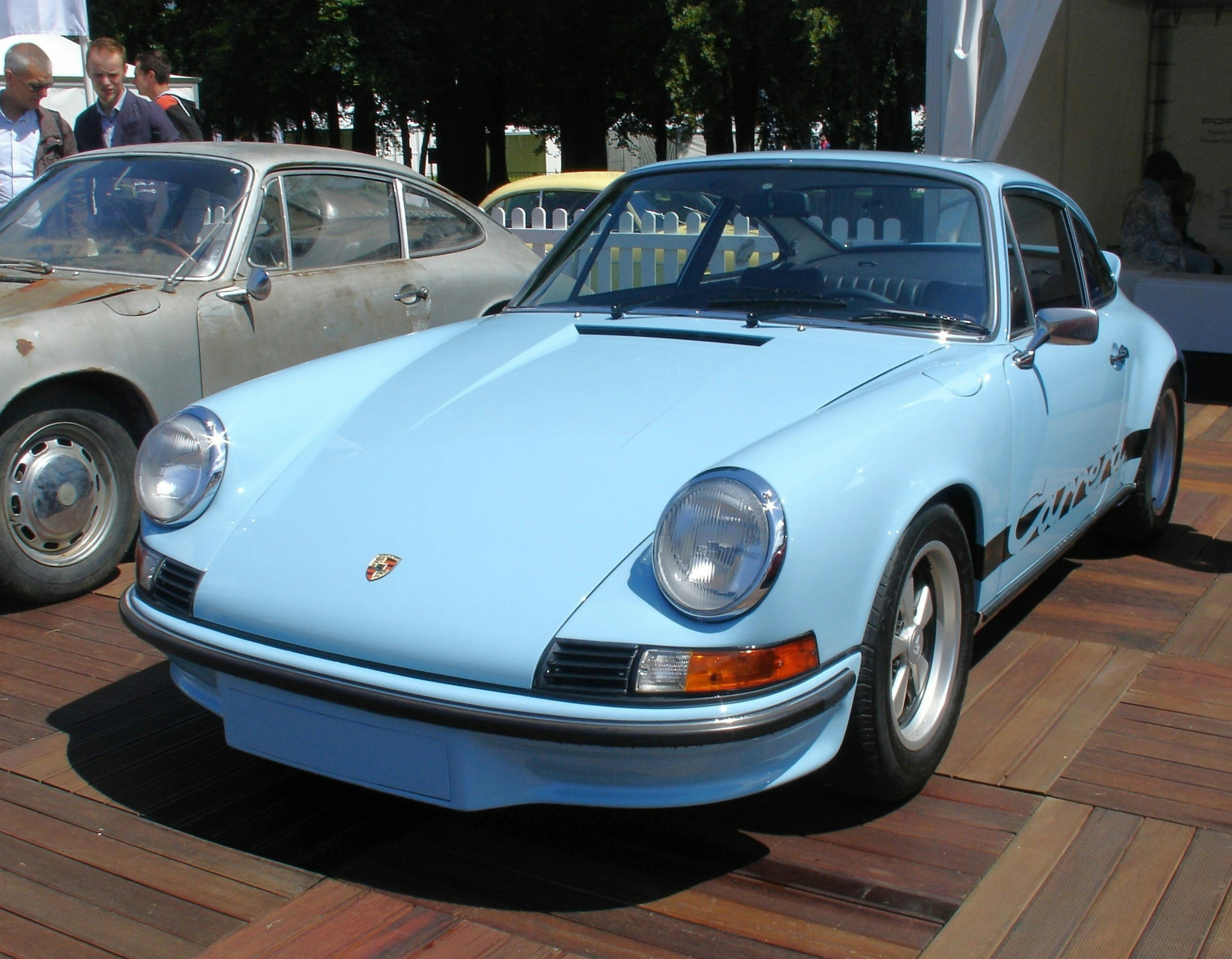 Blue Porsche 911 on a wooden platform at a car show, with vintage vehicles and onlookers in the background.