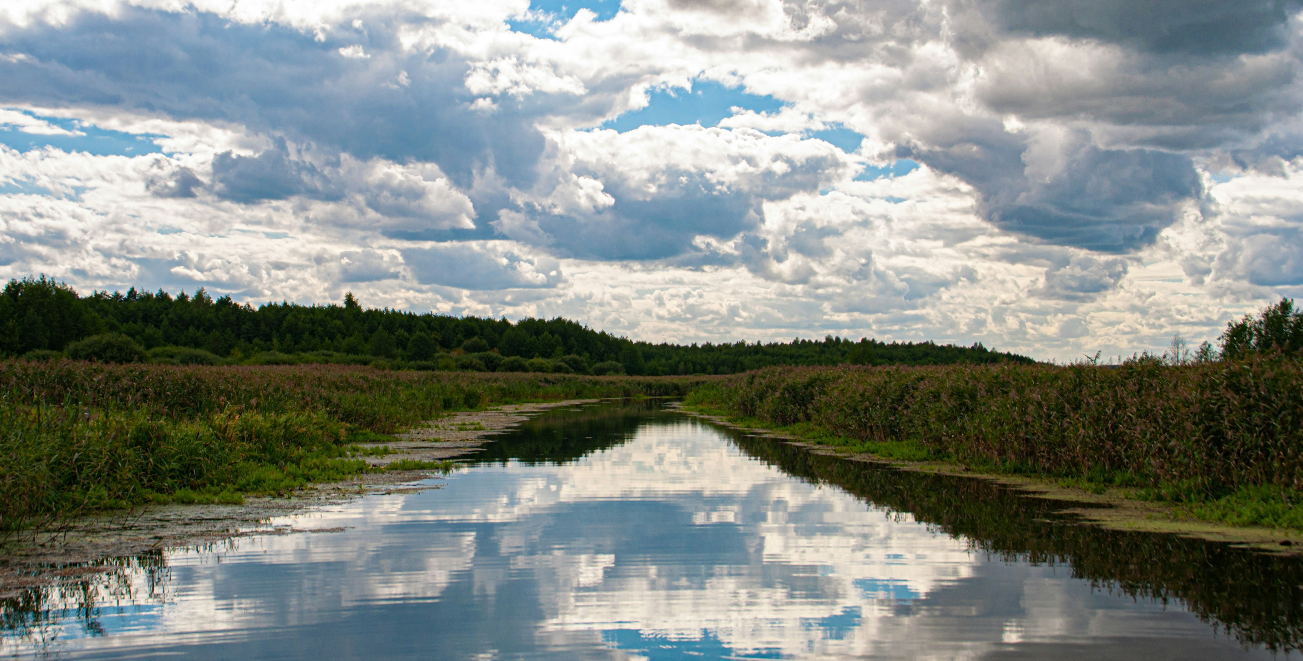 Wide wetland channel mirrors the sky as distant horizon and marsh grasses line a tranquil path, inviting exploration.