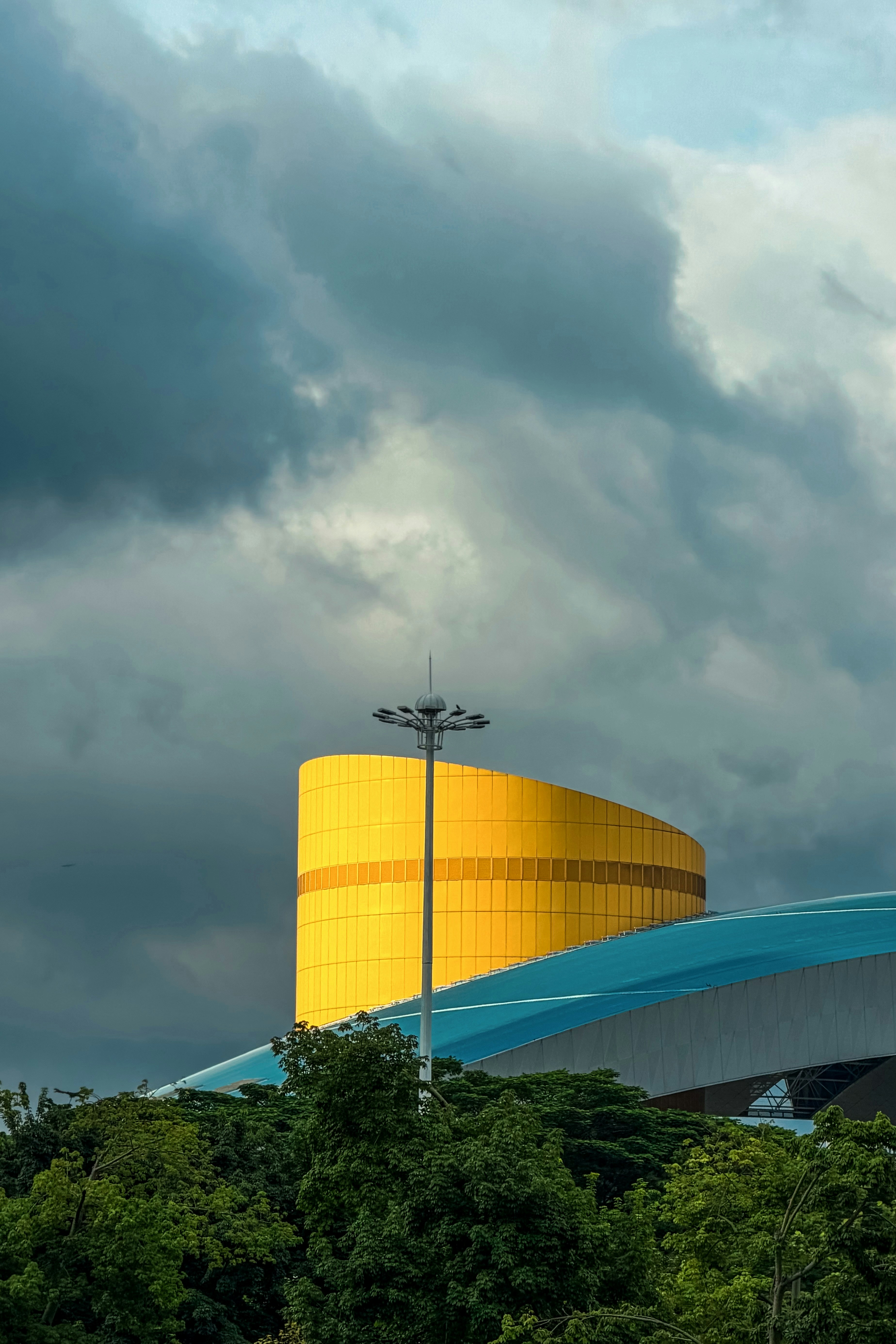 Architectural structure with a golden cylindrical facade peaks above a green treeline, under dramatic clouds. A modern roofline curves beneath, suggesting an urban landmark.