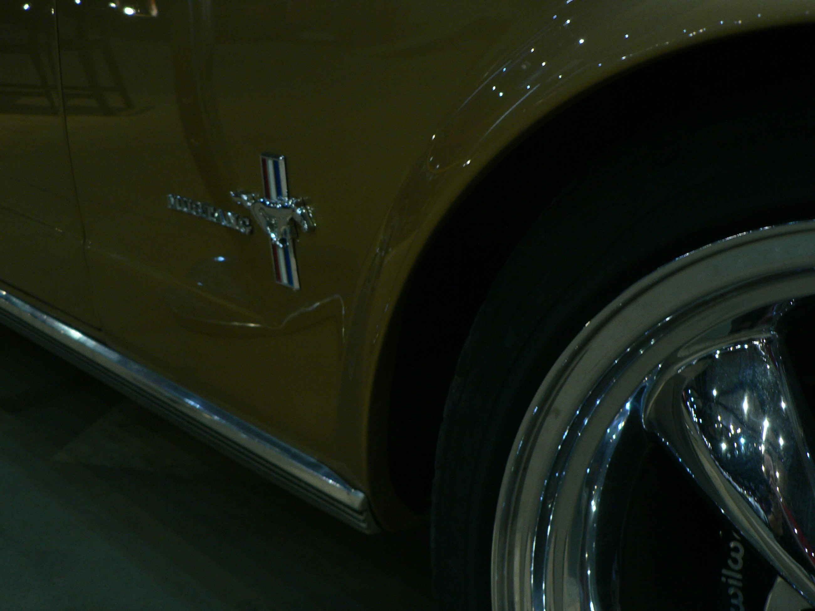 The image offers a close-up, moody shot of the fender and wheel well of a vintage Ford Mustang, rendered in a warm gold hue. The focal point is the iconic Mustang emblem—a galloping horse flanked by red, white, and blue stripes—mounted on the fender with polished chrome detailing. The bodywork’s glossy finish catches subtle highlights, emphasizing the car’s smooth curves. To the right, a portion of the wheel is visible: a highly polished chrome rim reflects ambient light, while the black tire contrasts sharply with the metallic sheen.
