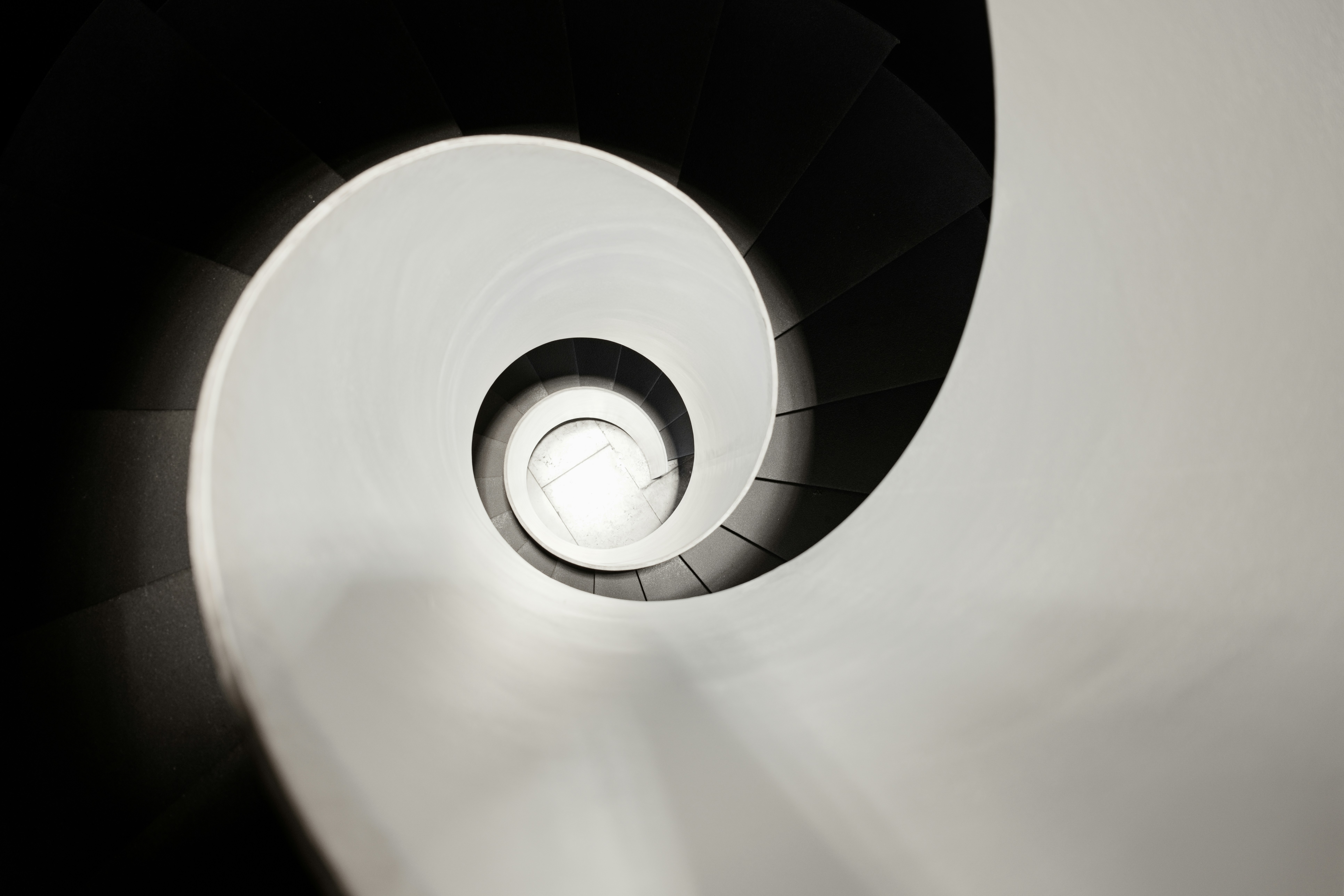 Monochrome spiral staircase viewed from above, drawing the eye toward the central lantern. The shot emphasizes curves and depth with soft shading and smooth tonal transitions.