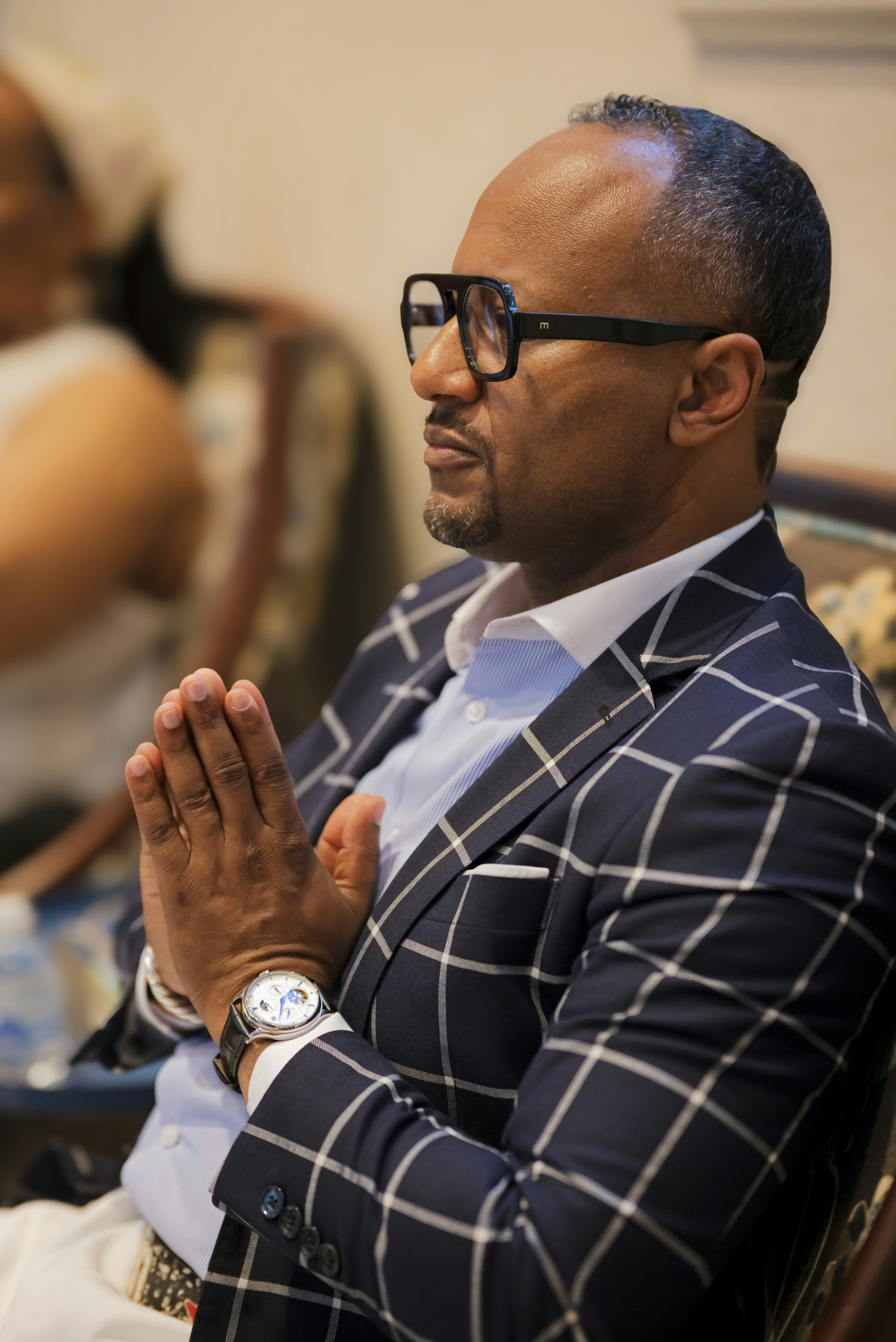 A well-dressed man in a checkered blazer sits in profile with hands in prayer. The composition centers on his face, glasses, and folded hands, suggesting a moment of contemplation at an event. The scene is candid and polished.