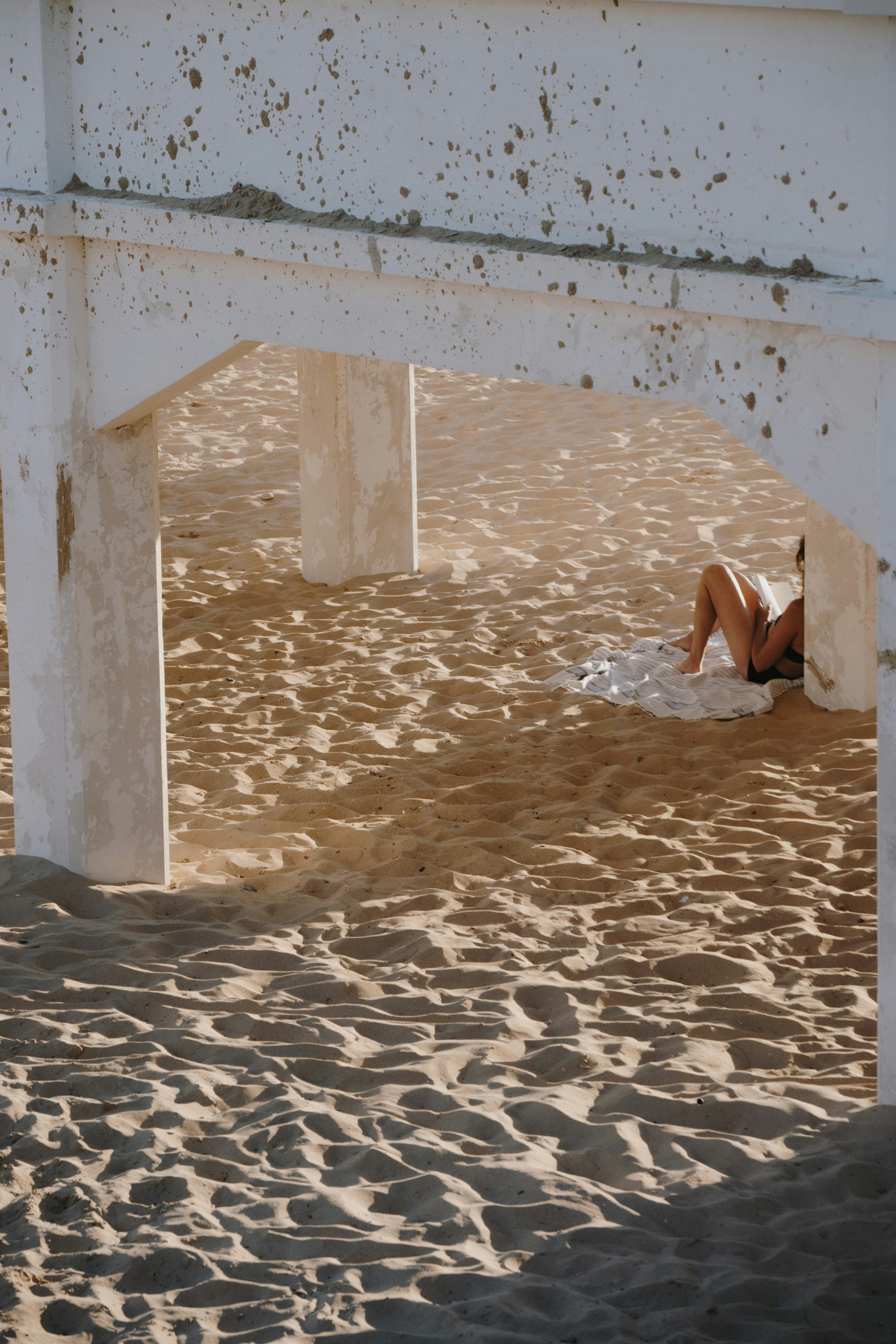 Shade beneath a weathered pier: sunlit sand and a lone swimmer