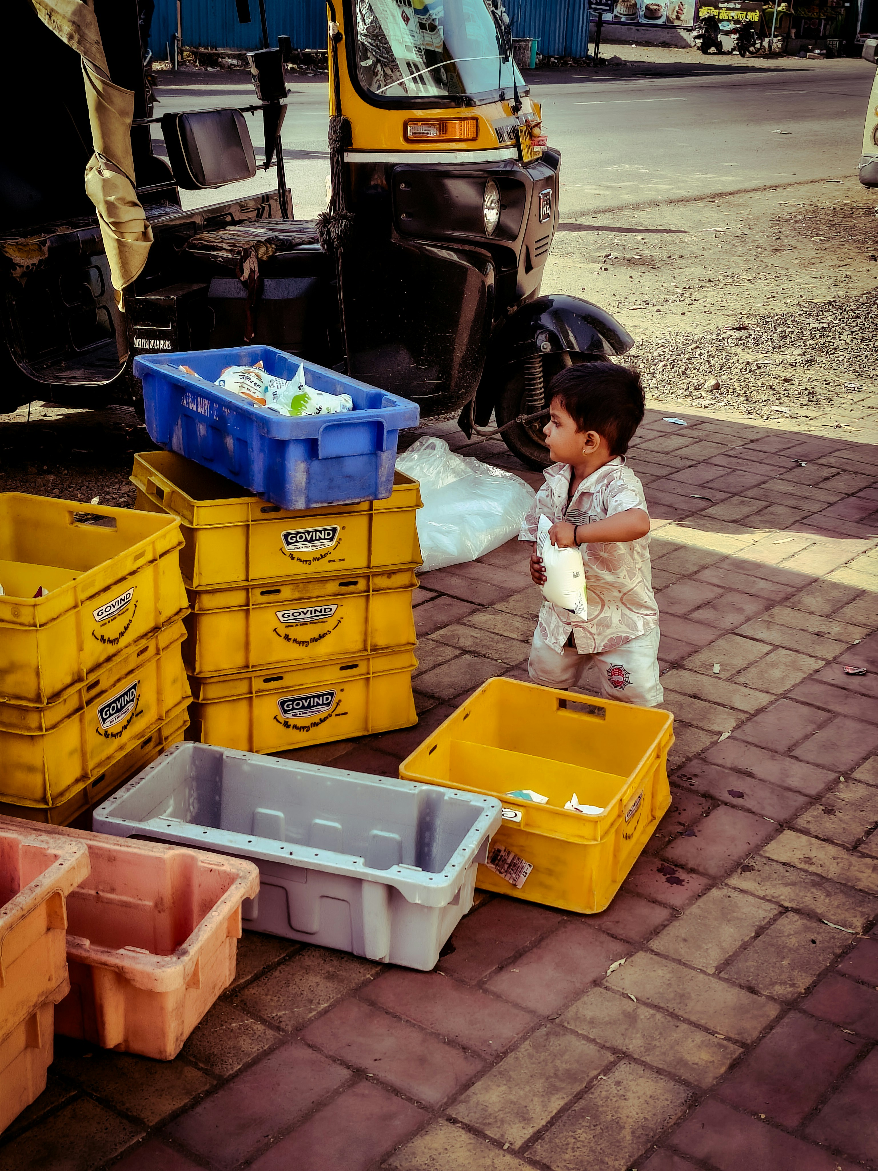 A child holding a plastic milk pouch beside stacked yellow crates and a rickshaw
