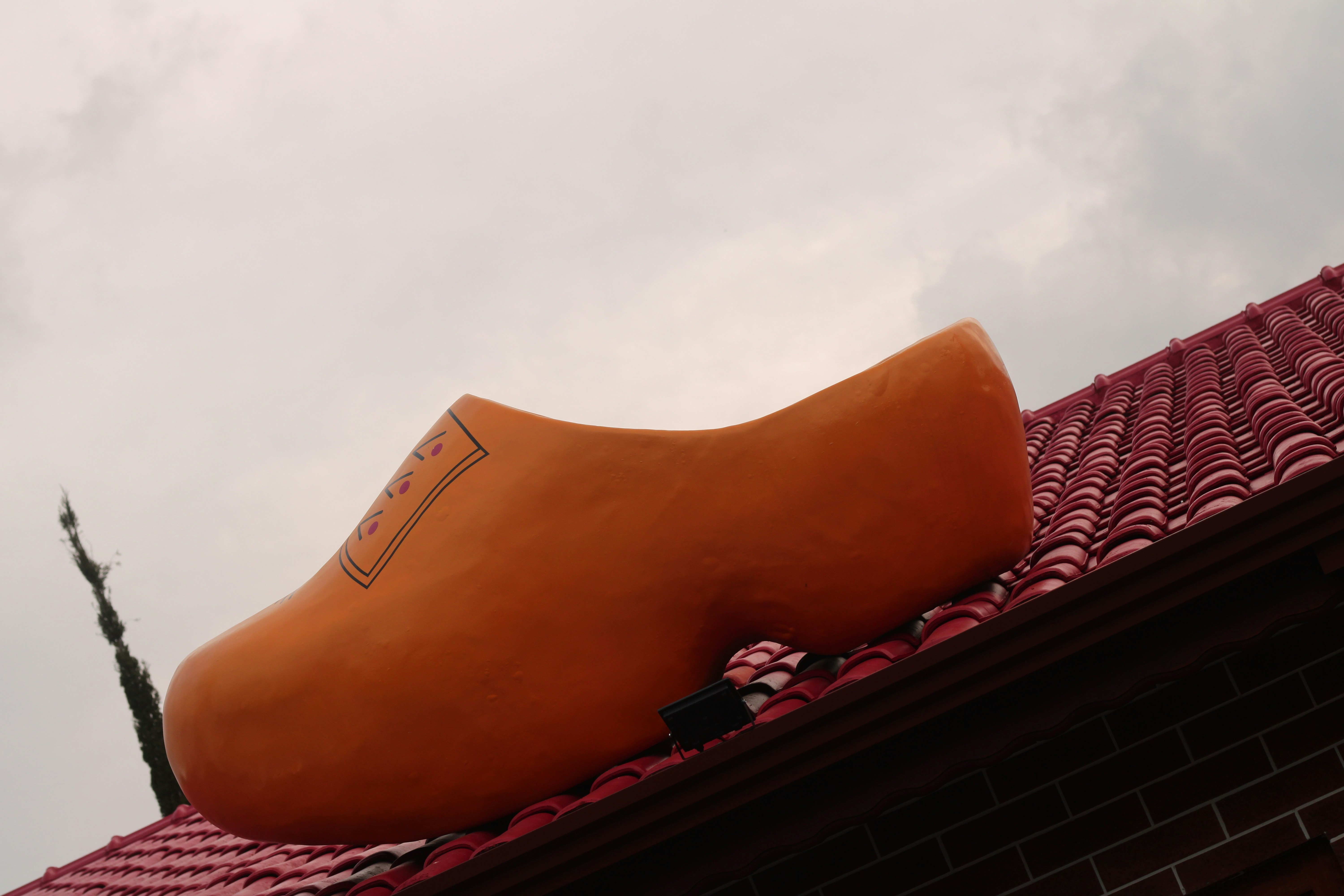 A massive orange shoe sculpture perched on a red-tiled roof against a muted sky. The scene highlights the whimsical, oversized footwear in an architectural setting.