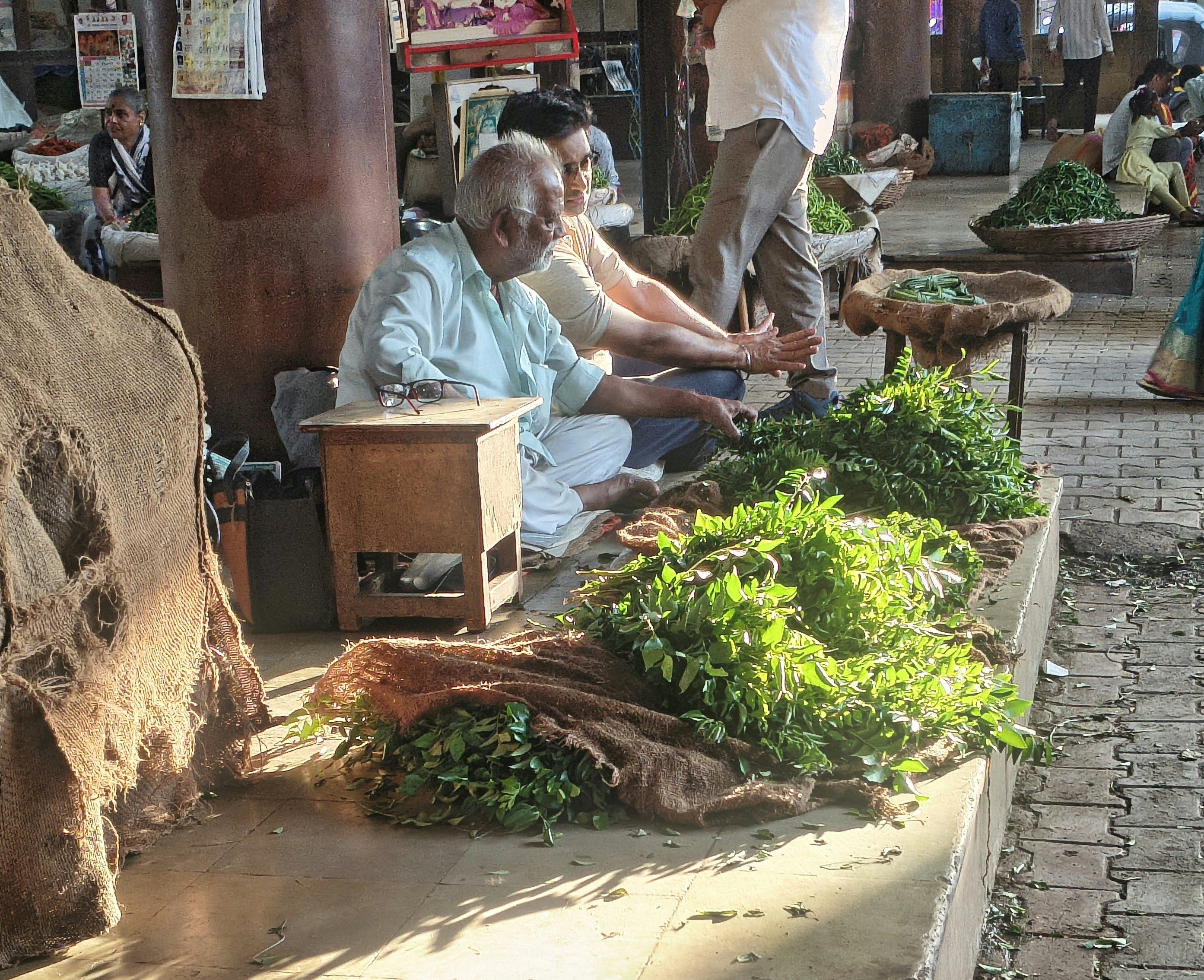Market scene: elder man and helper arranging fresh greens by a busy stall