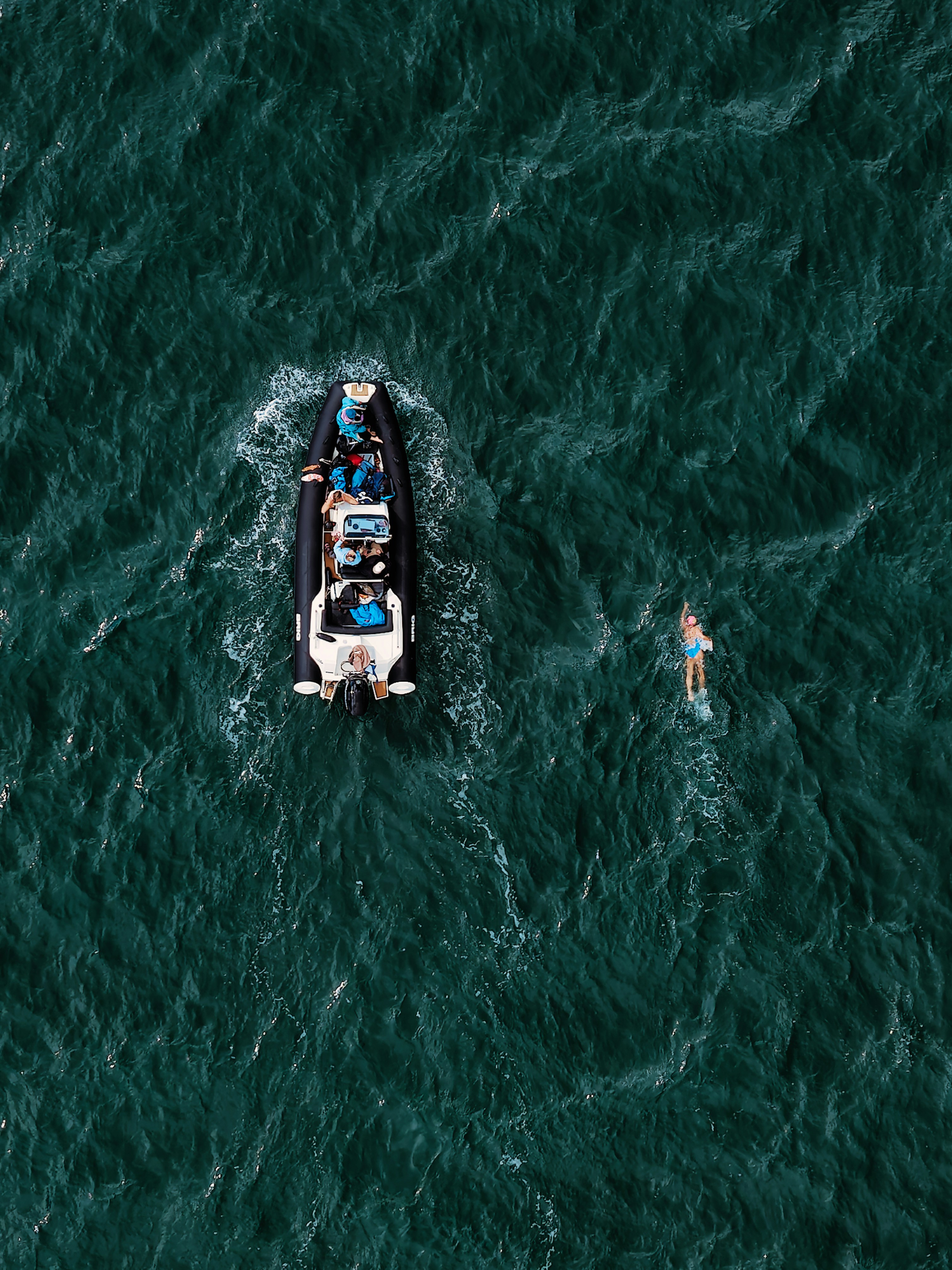 Aerial view of a small boat with several passengers cruising through teal sea, a swimmer nearby, all captured from above to emphasize motion and water texture.