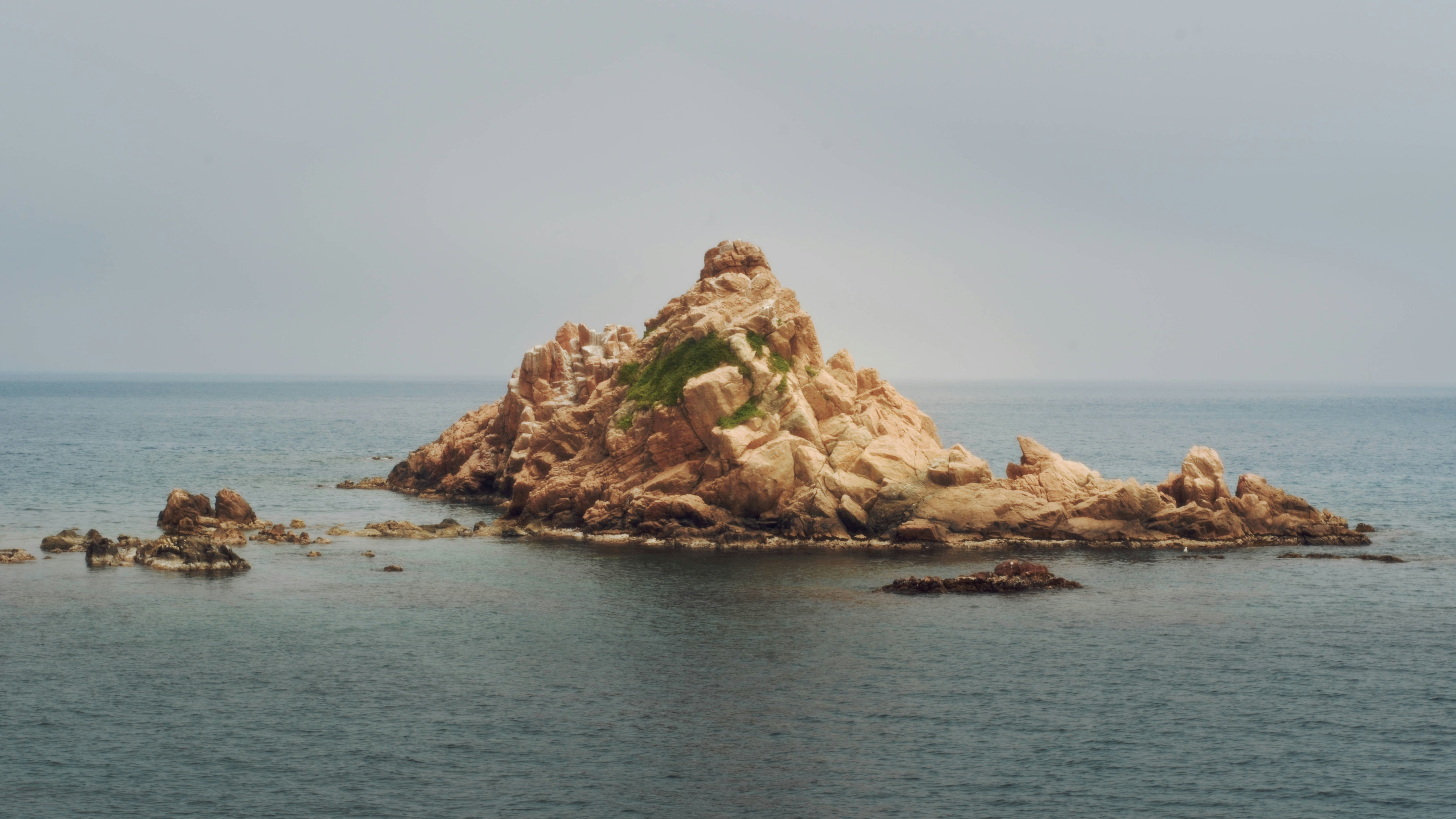 A solitary rocky islet rises from calm sea waters under a hazy sky, with sparse green growth on its rugged surface. The scene emphasizes isolation and natural geology without human presence.