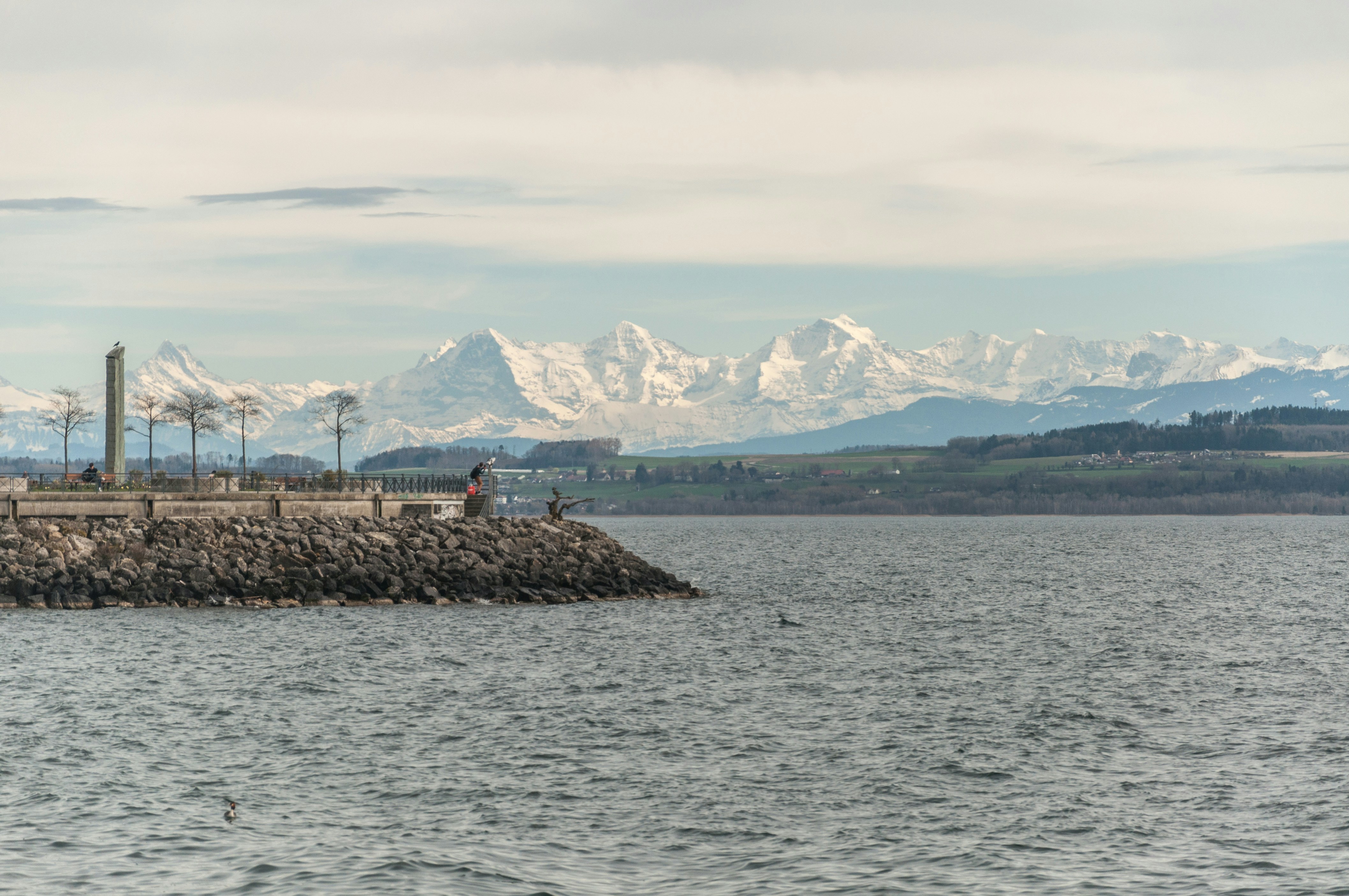 Calm sea with a rocky breakwater extending into the water; distant snow-capped mountains rise under a pale sky.