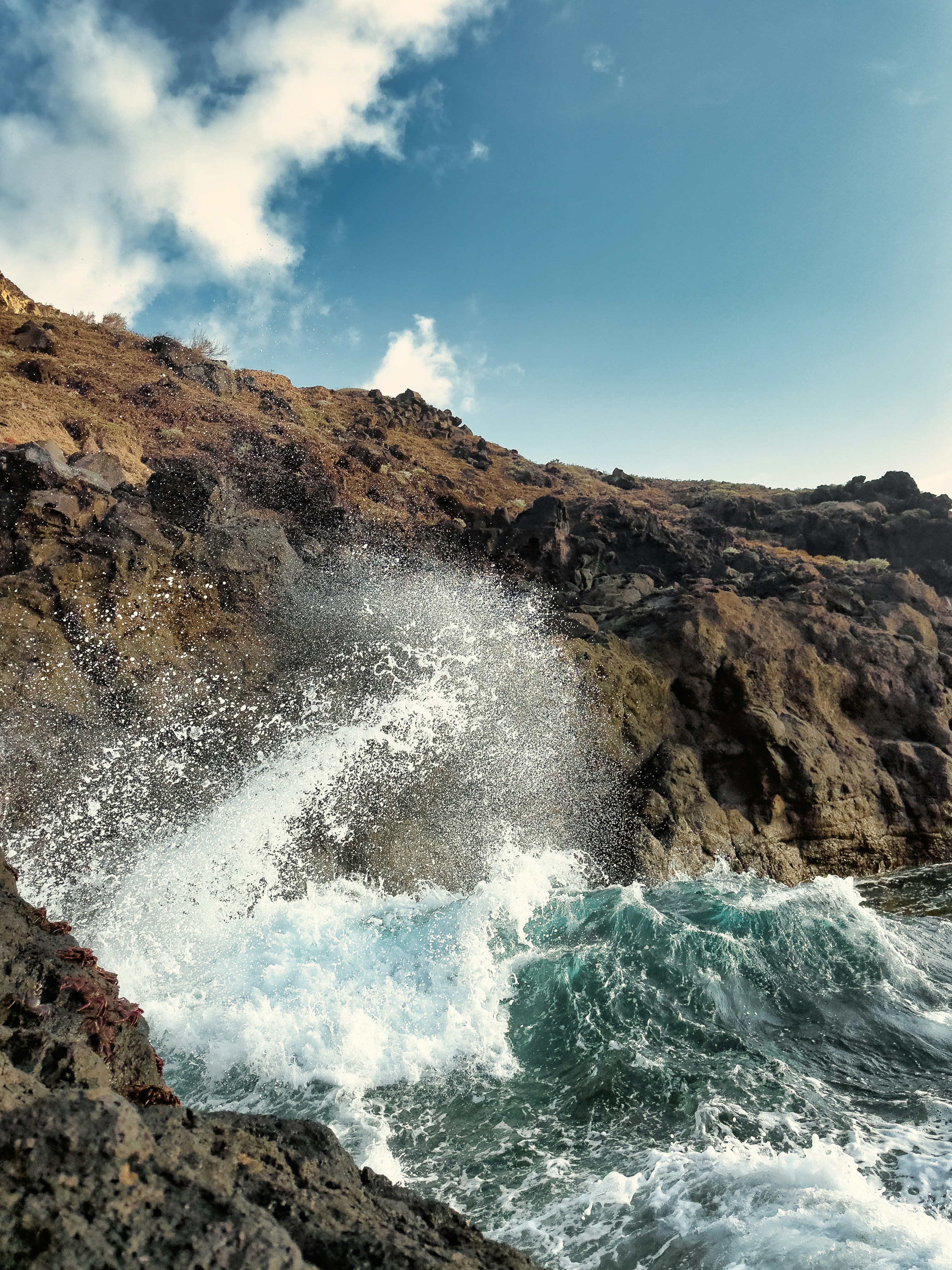 Crashing turquoise waves collide with jagged volcanic rocks, sending a spray plume over a rugged shoreline under a bright blue sky.