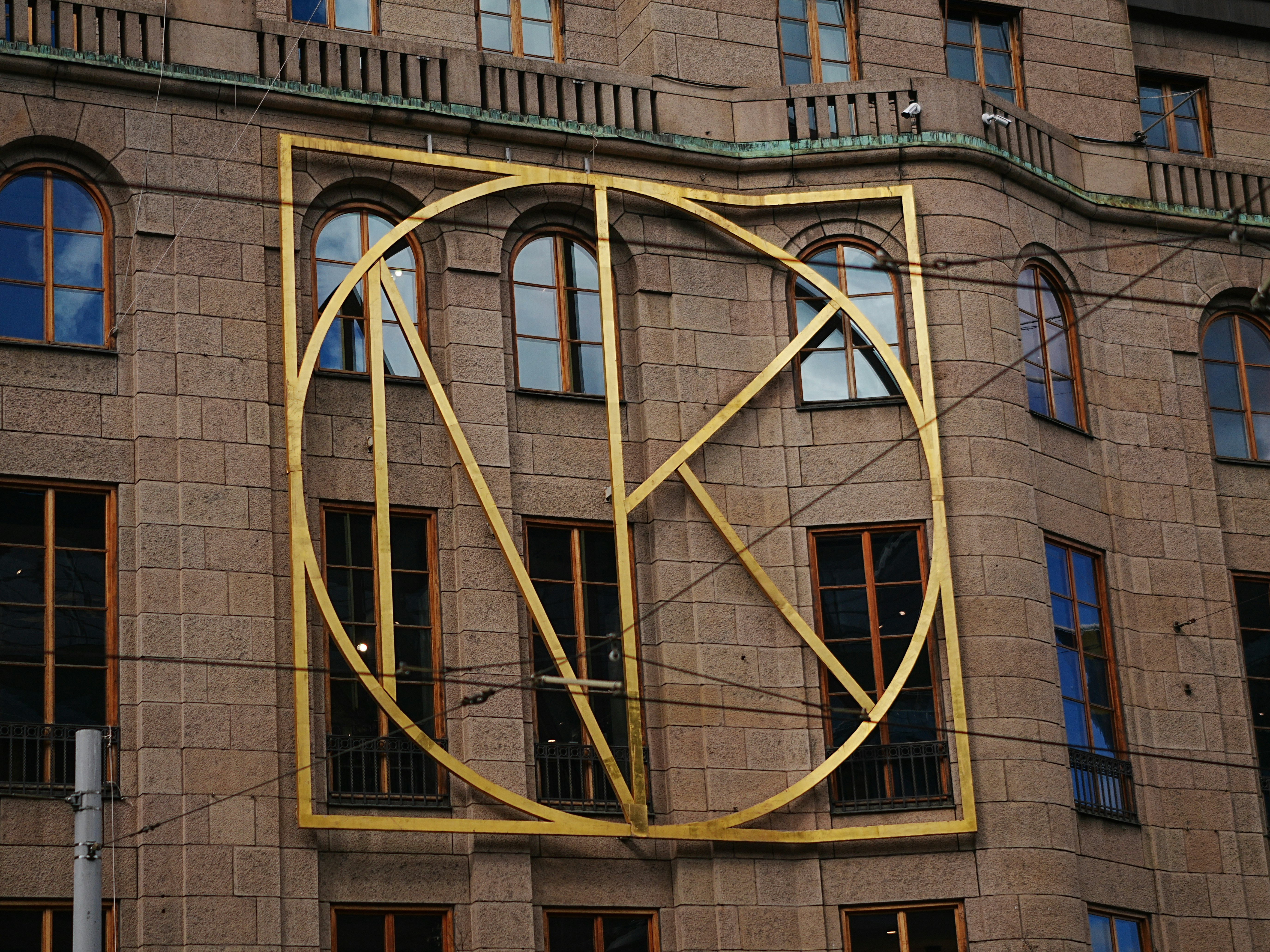 A large gold geometric sculpture mounted on a stone building facade with arched windows