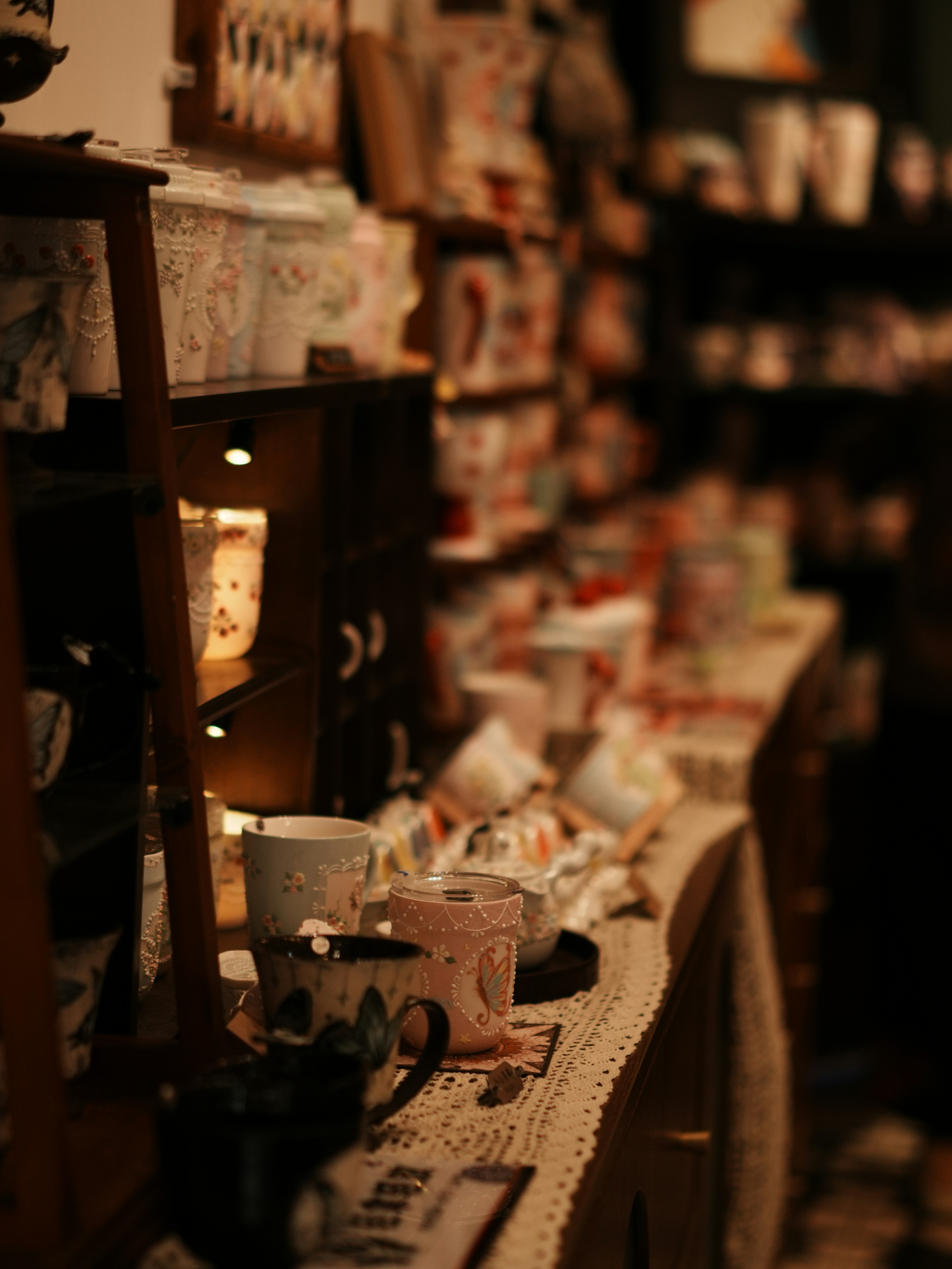 Rows of decorative mugs and cups line a rustic counter in a cozy shop, with warm ambient lighting highlighting their patterns.