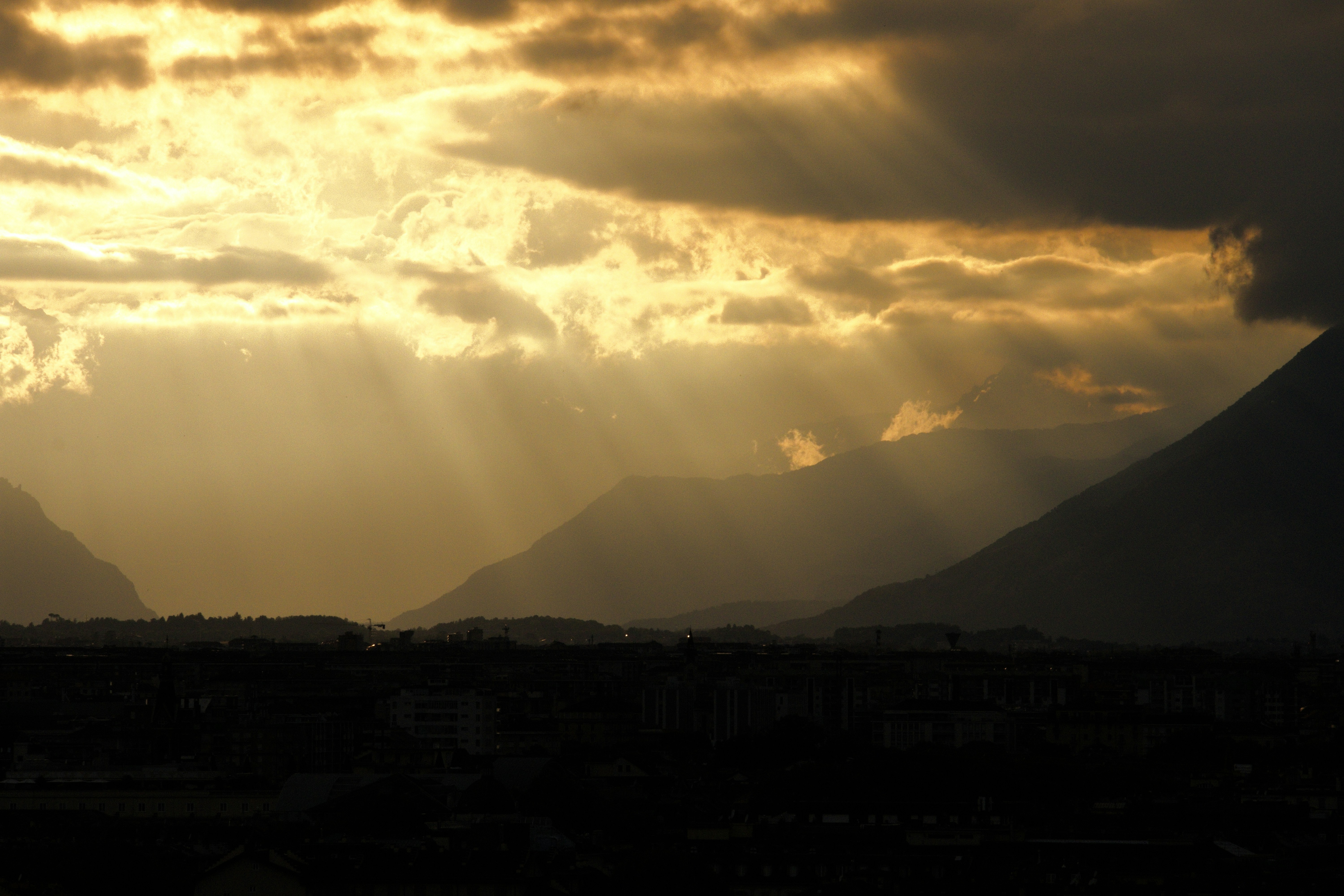 Golden sunrays pierce a hazy sky over a silhouetted city with distant mountains; dramatic light creates a calm, moody horizon.
