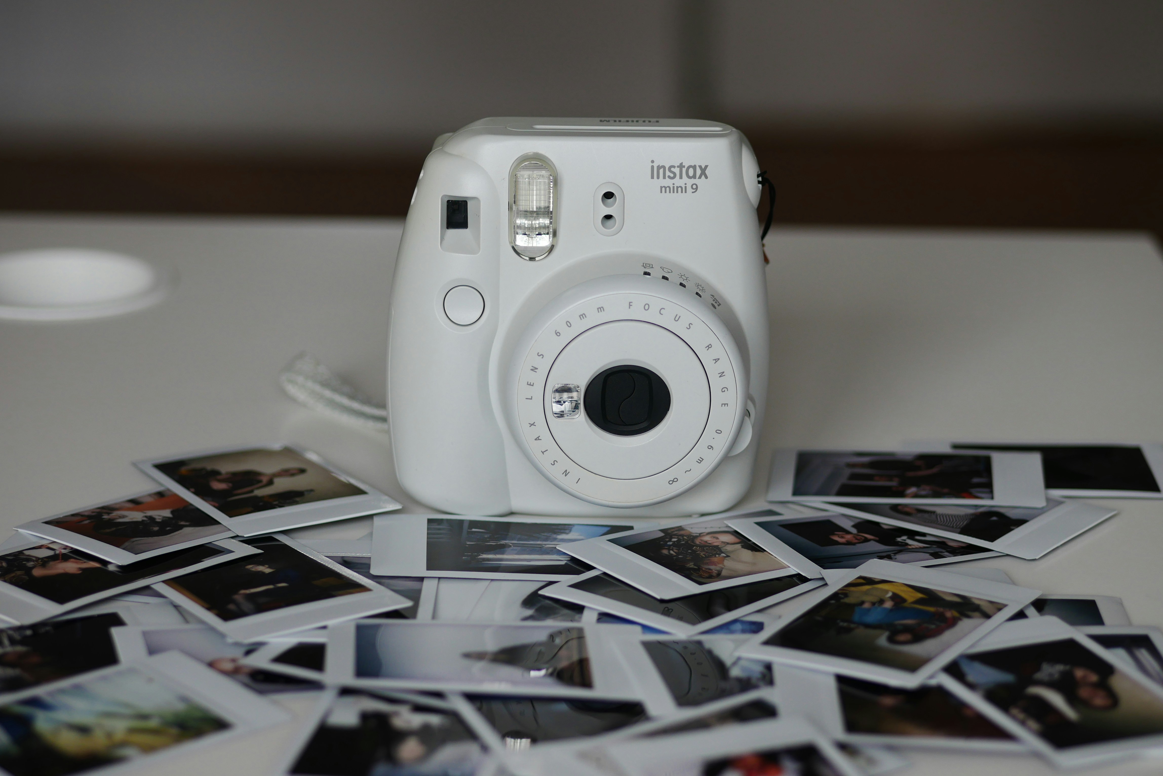 White Instax mini 9 sits among scattered photo prints on a tabletop, the quirky compact camera serving as the focal point of a casual still-life scene.