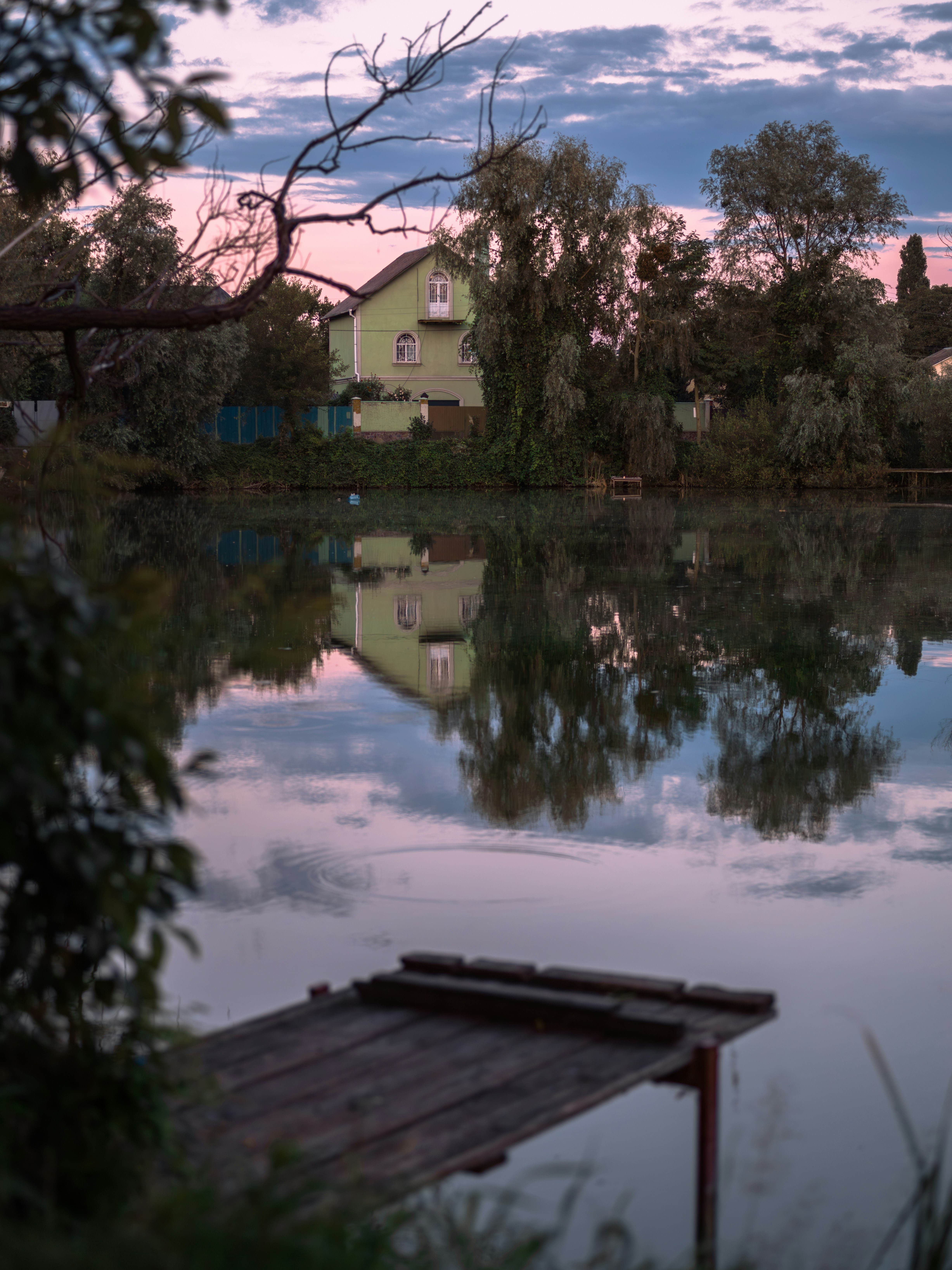 A green house reflected in a calm lake with a wooden pier in foreground