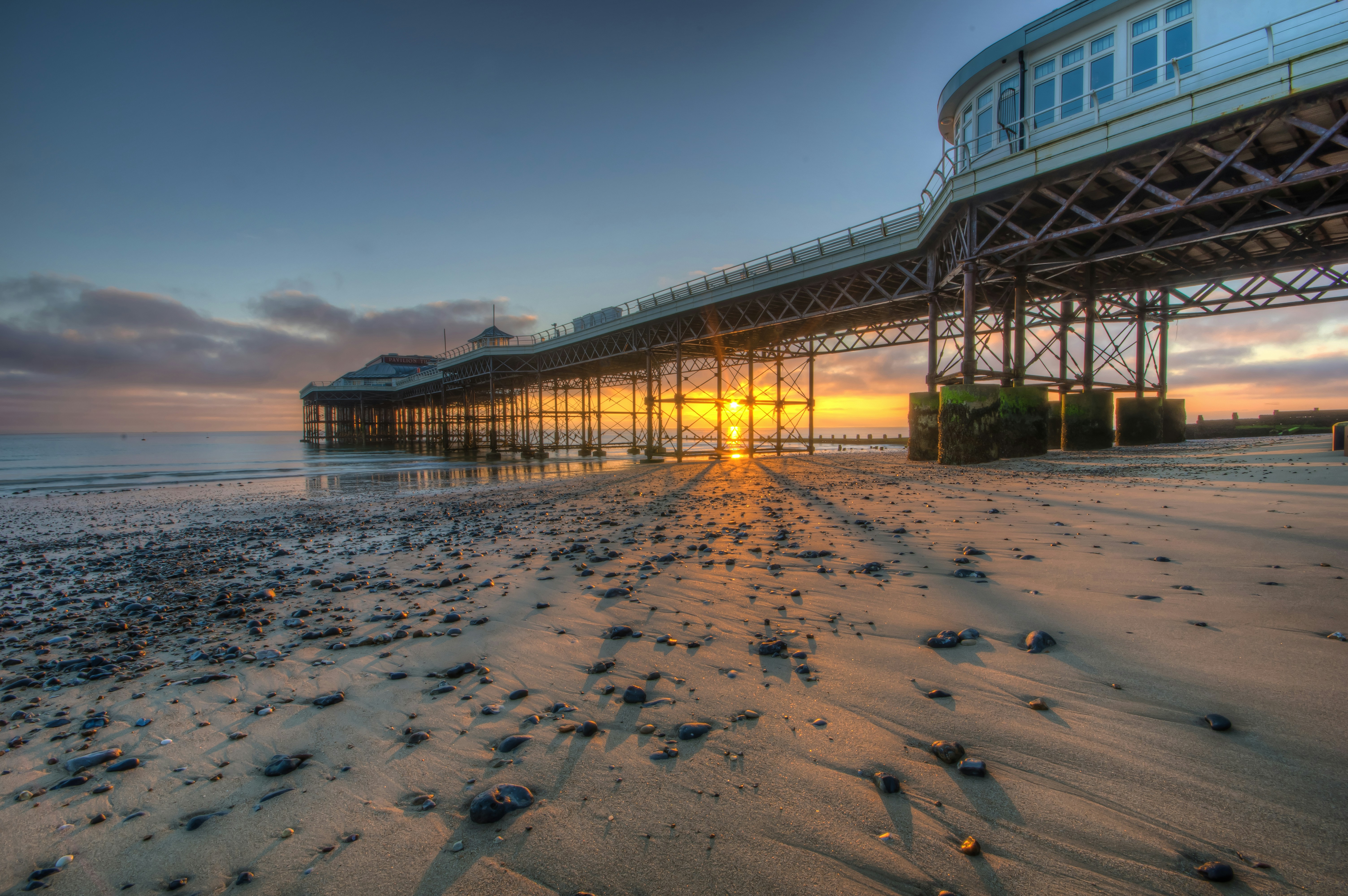 Sunset glows over a pier extending into calm water, with wet sand and scattered shells in the foreground. The scene emphasizes the structural lines of the pier and the warm horizon at golden hour.