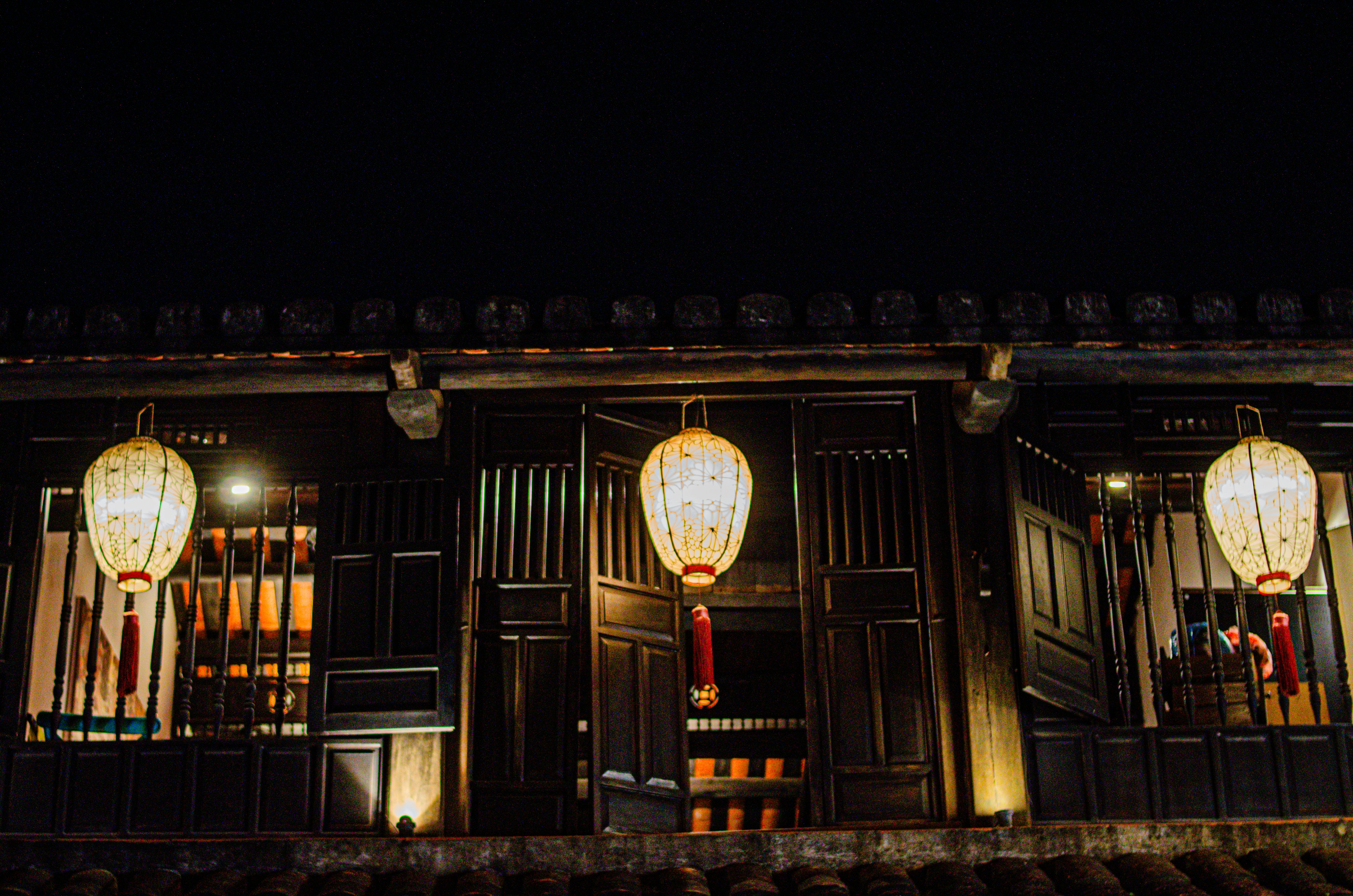 Red lanterns illuminate a dark temple corridor with open wooden doors and warm interior glow.
