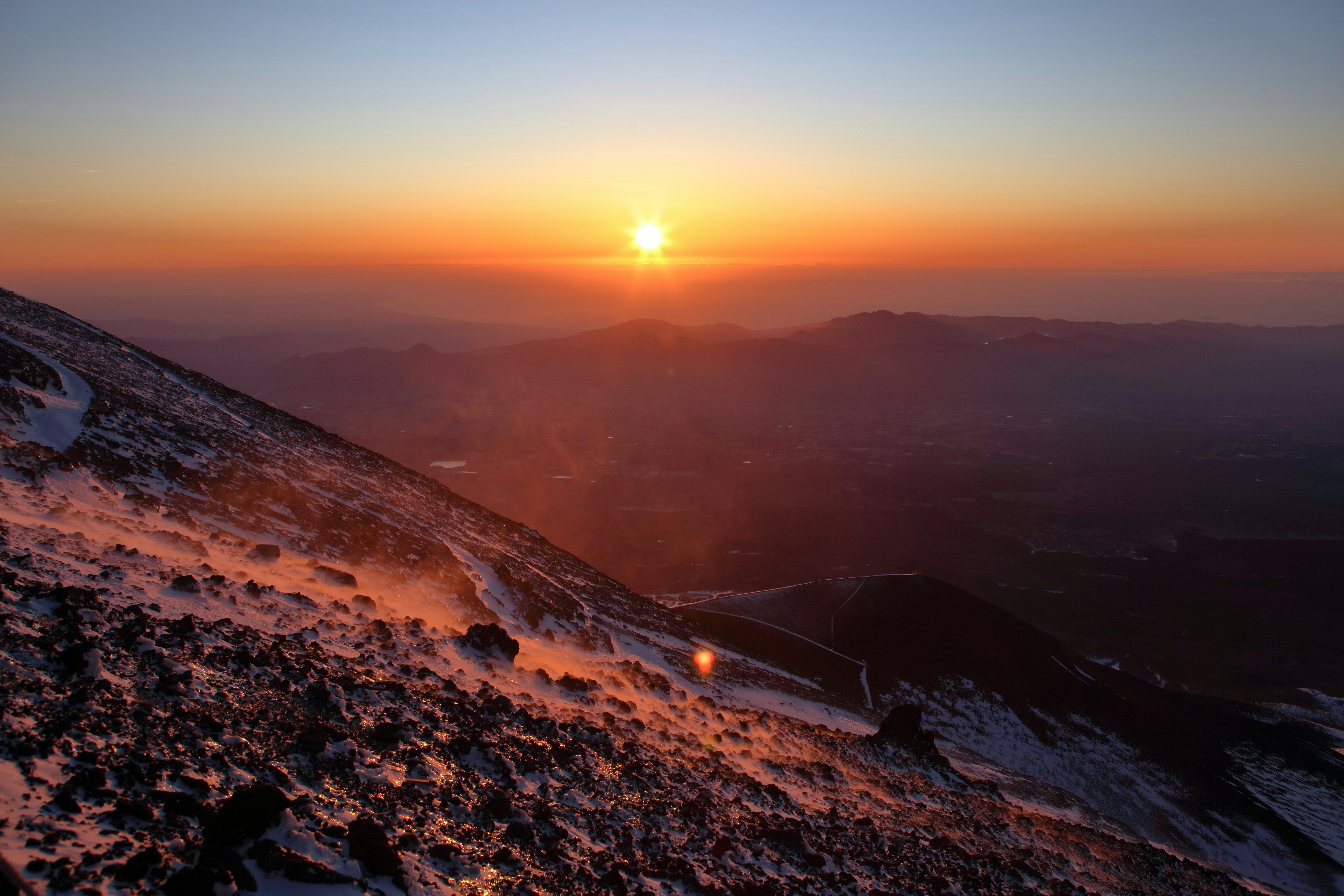 Sunset casts warm hues over snow-dusted slopes and a distant, hazy valley, with a winding path and a dark ridge guiding the eye toward the radiant sun.