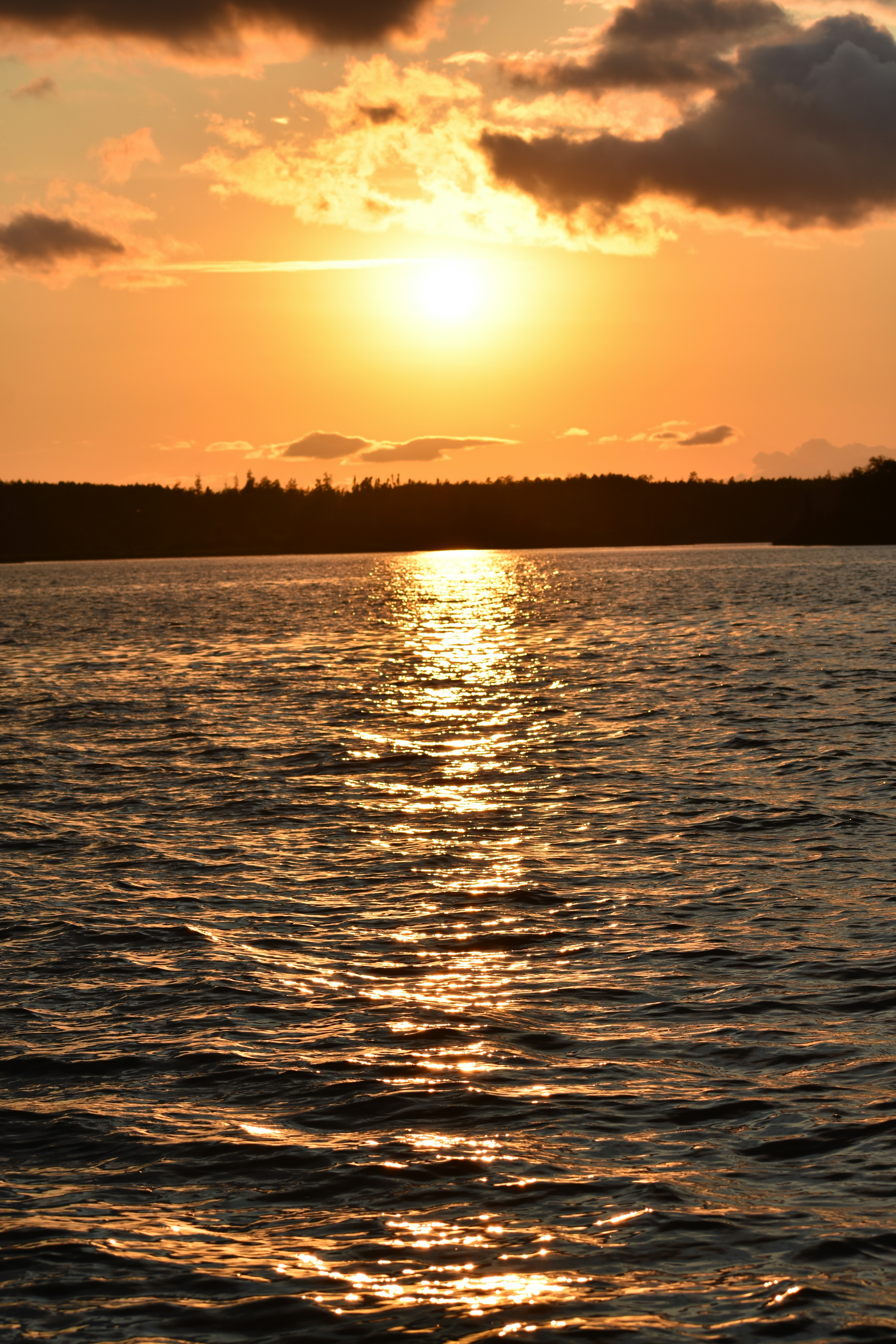 Rich orange sunset over rippling water with a dark distant shoreline; minimal clouds and a bright sun casting a reflective path on the lake.