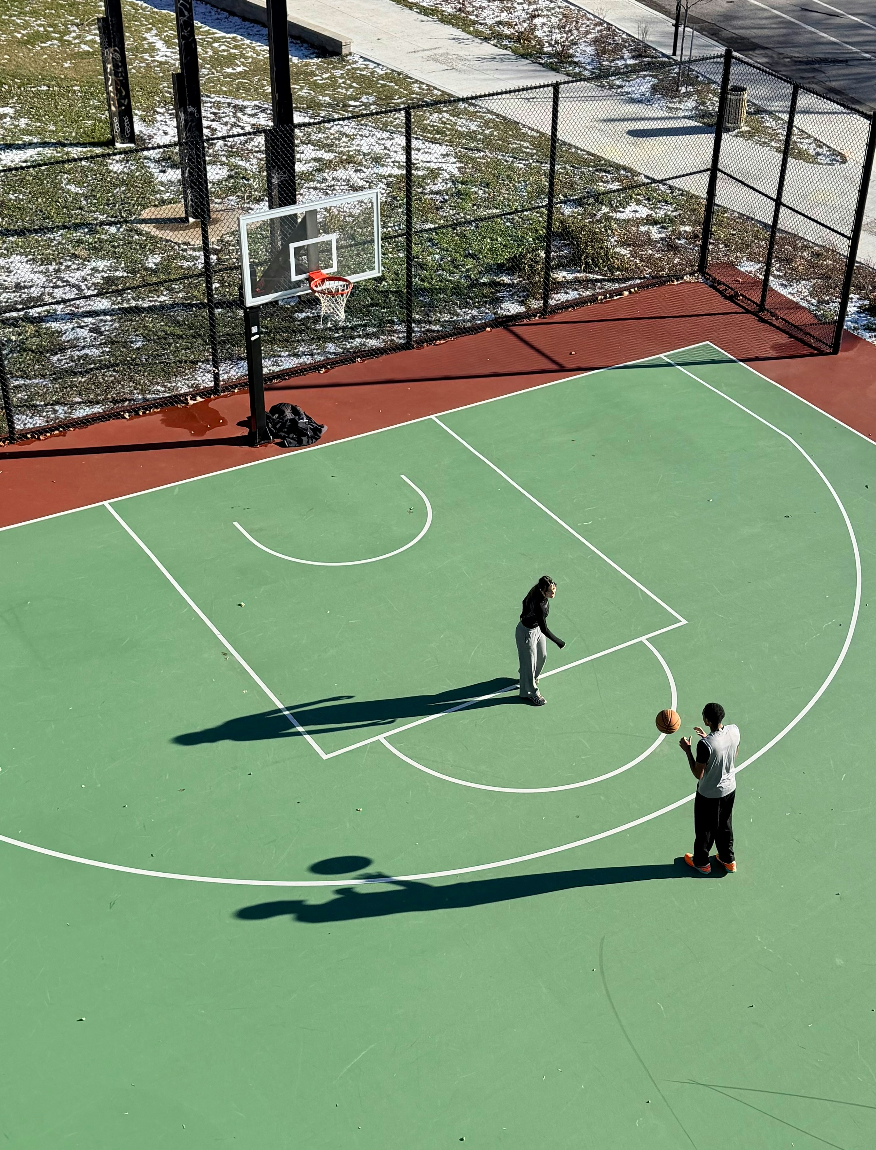Two players on a green outdoor basketball court with a hoop, long shadows, and a fence surrounding the court.