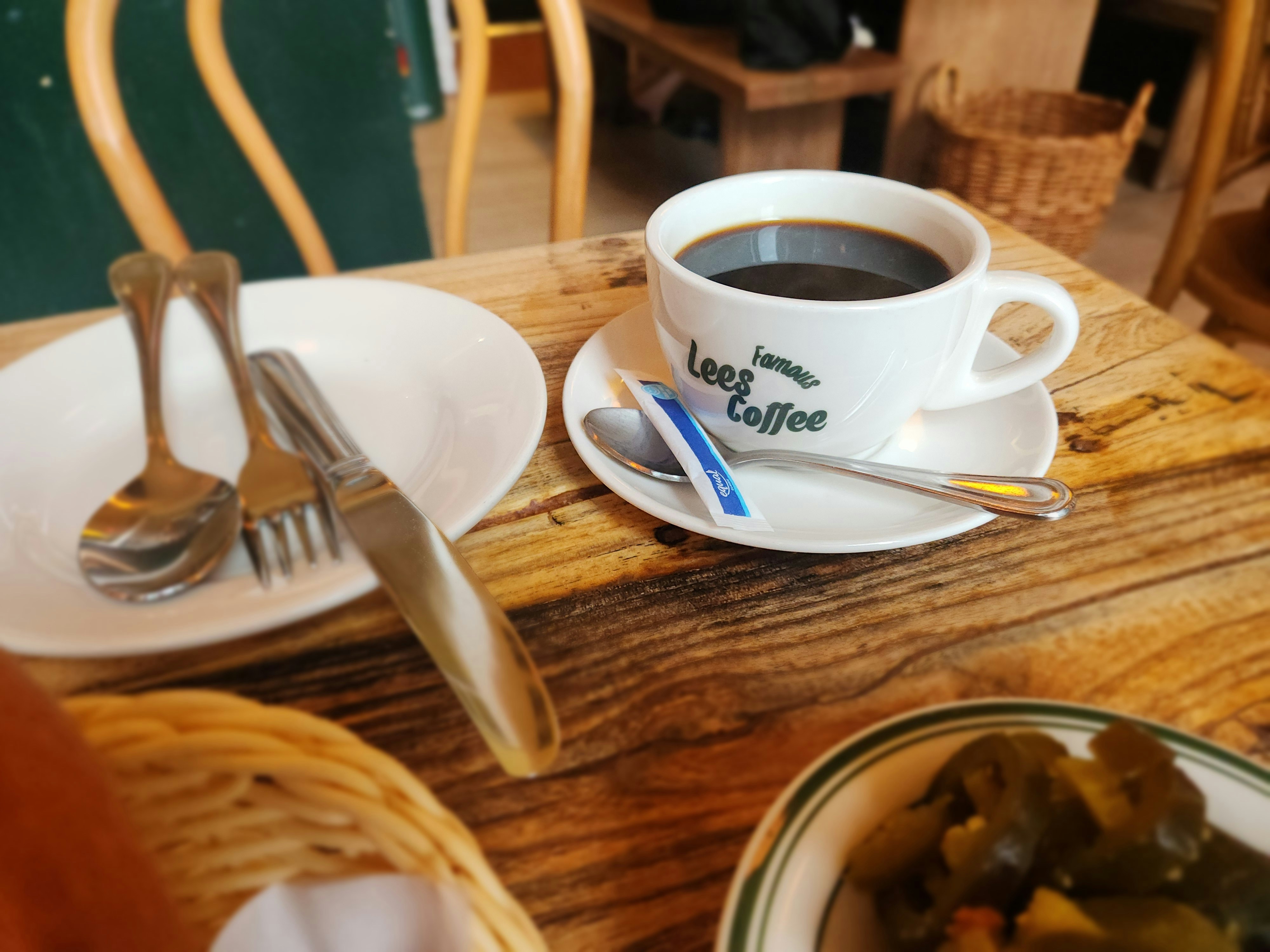 A cup of coffee on a saucer sits on a wooden table beside a basket and cutlery, with another dish partially visible; warm cafe scene emphasizing texture and everyday ritual.